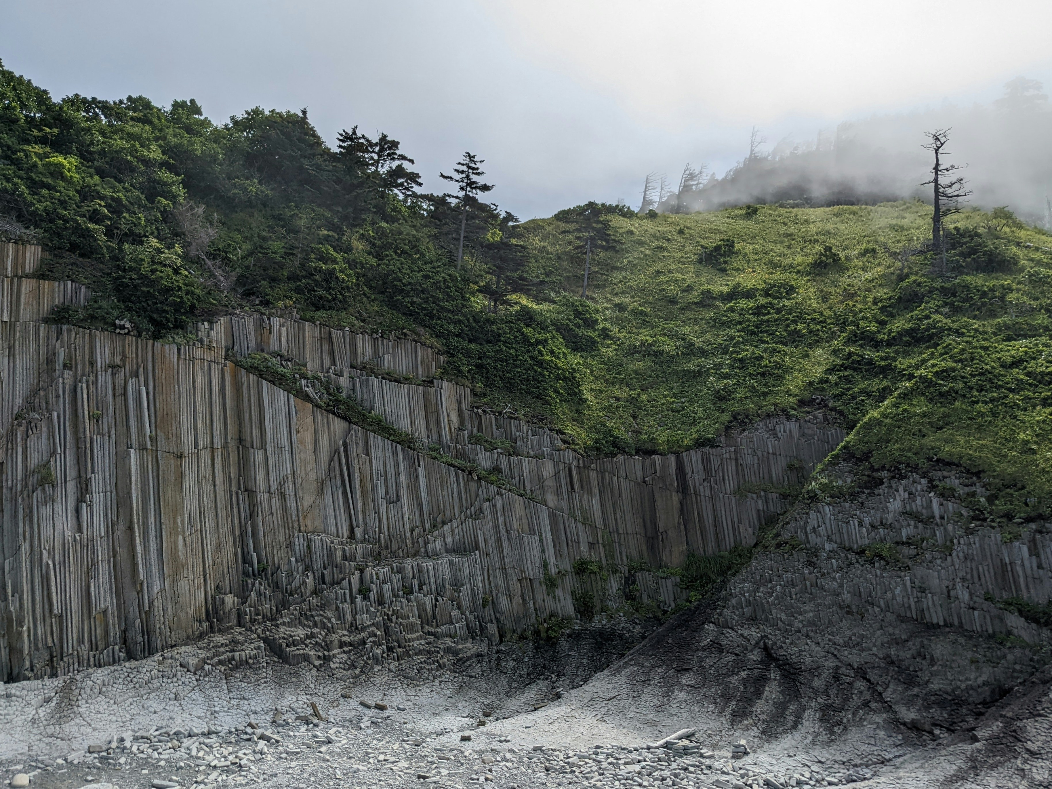Unique rock formations rise dramatically from the shoreline, framed by lush greenery and misty skies. The interplay of textures and colors highlights the geological history of the landscape.