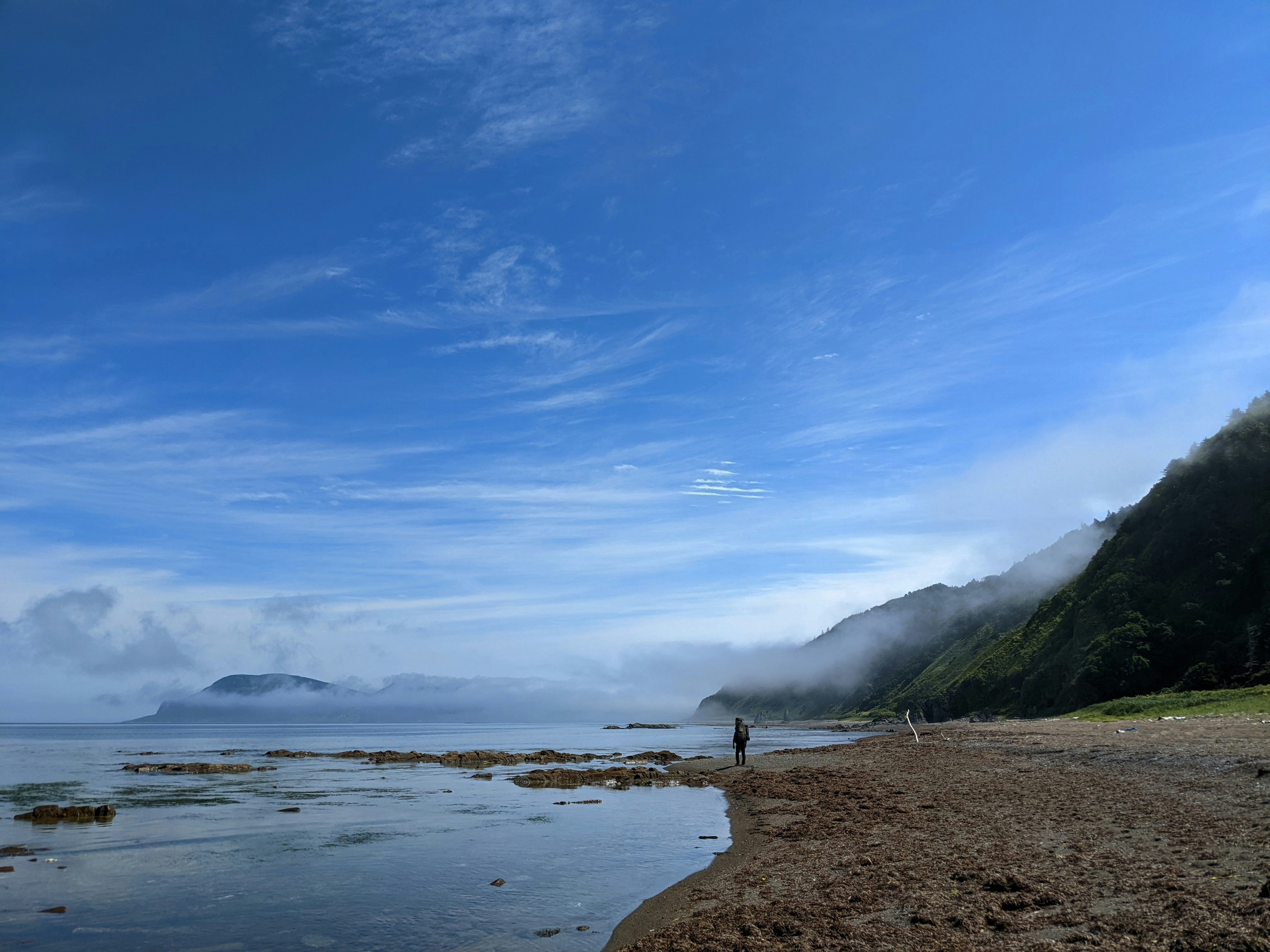 A lone figure walks along a serene beach, with misty hills and a calm sea under a vast blue sky. The scene captures the tranquil essence of coastal life.