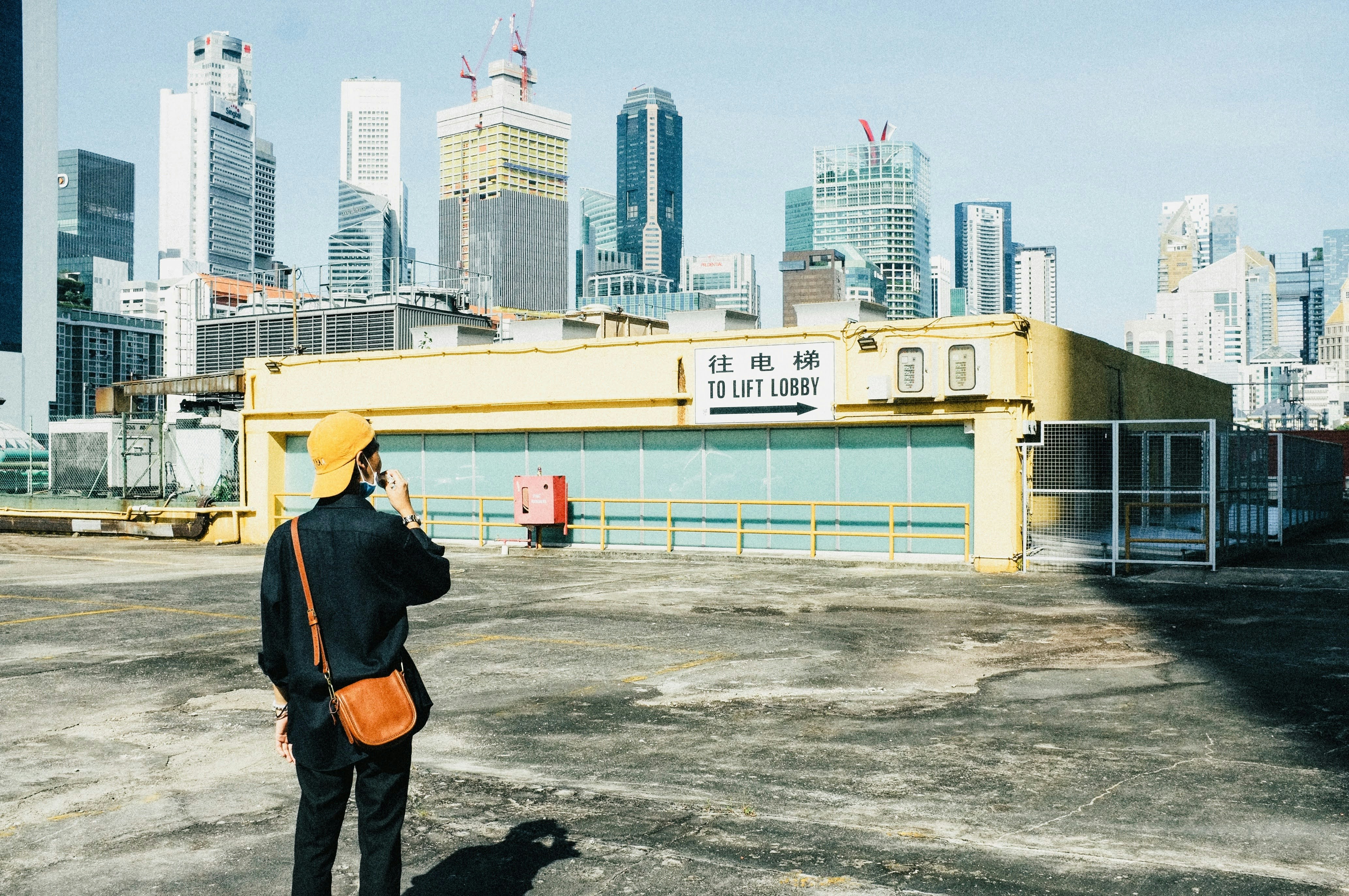 A person in casual attire stands on a rooftop, engaged in a phone call, with a backdrop of towering skyscrapers and a vibrant yellow building marked 'TO LIFT LOBBY.'