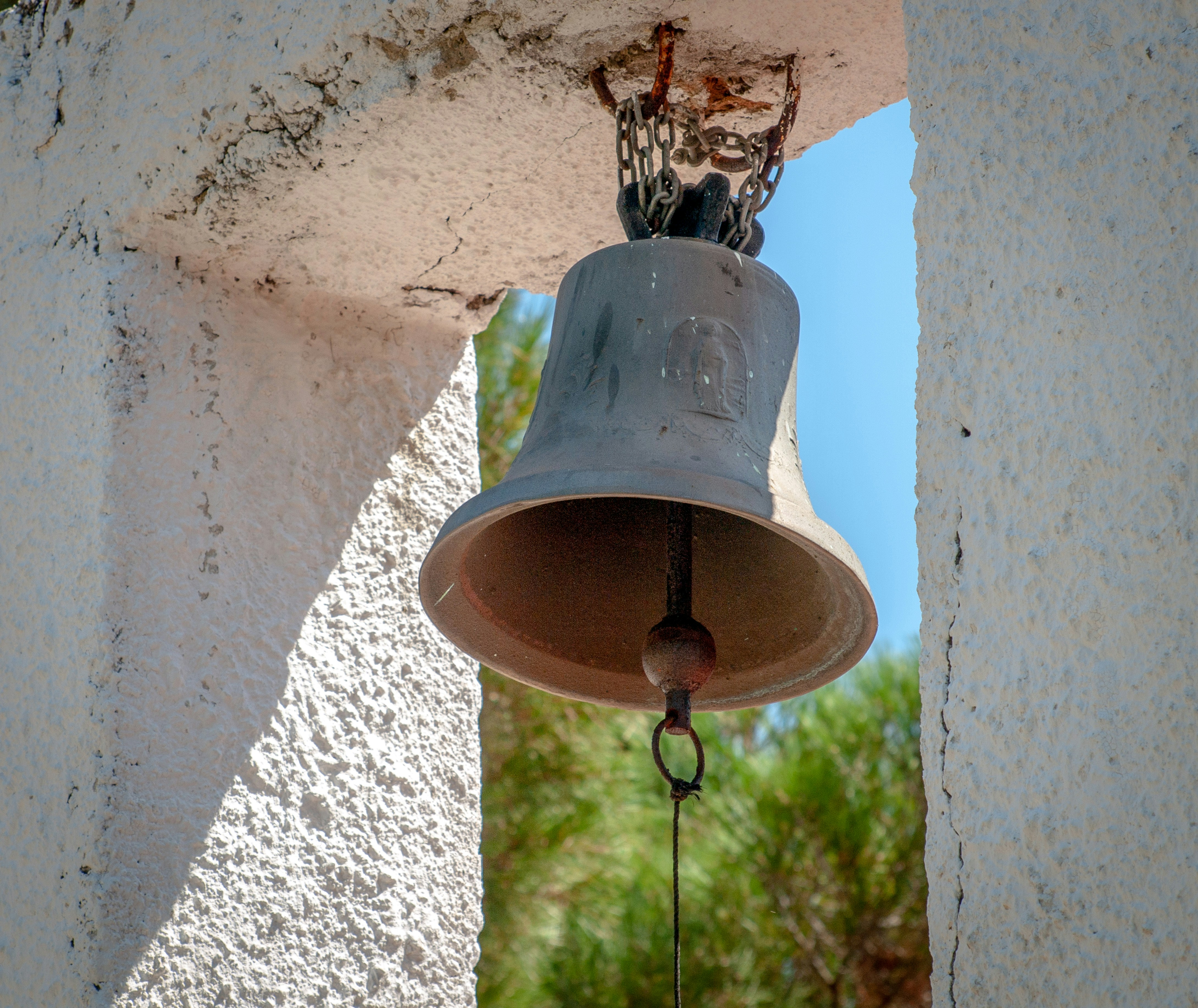 A weathered bell hangs from a rustic frame, surrounded by textured white walls and hints of greenery. Its silent presence evokes a sense of history.