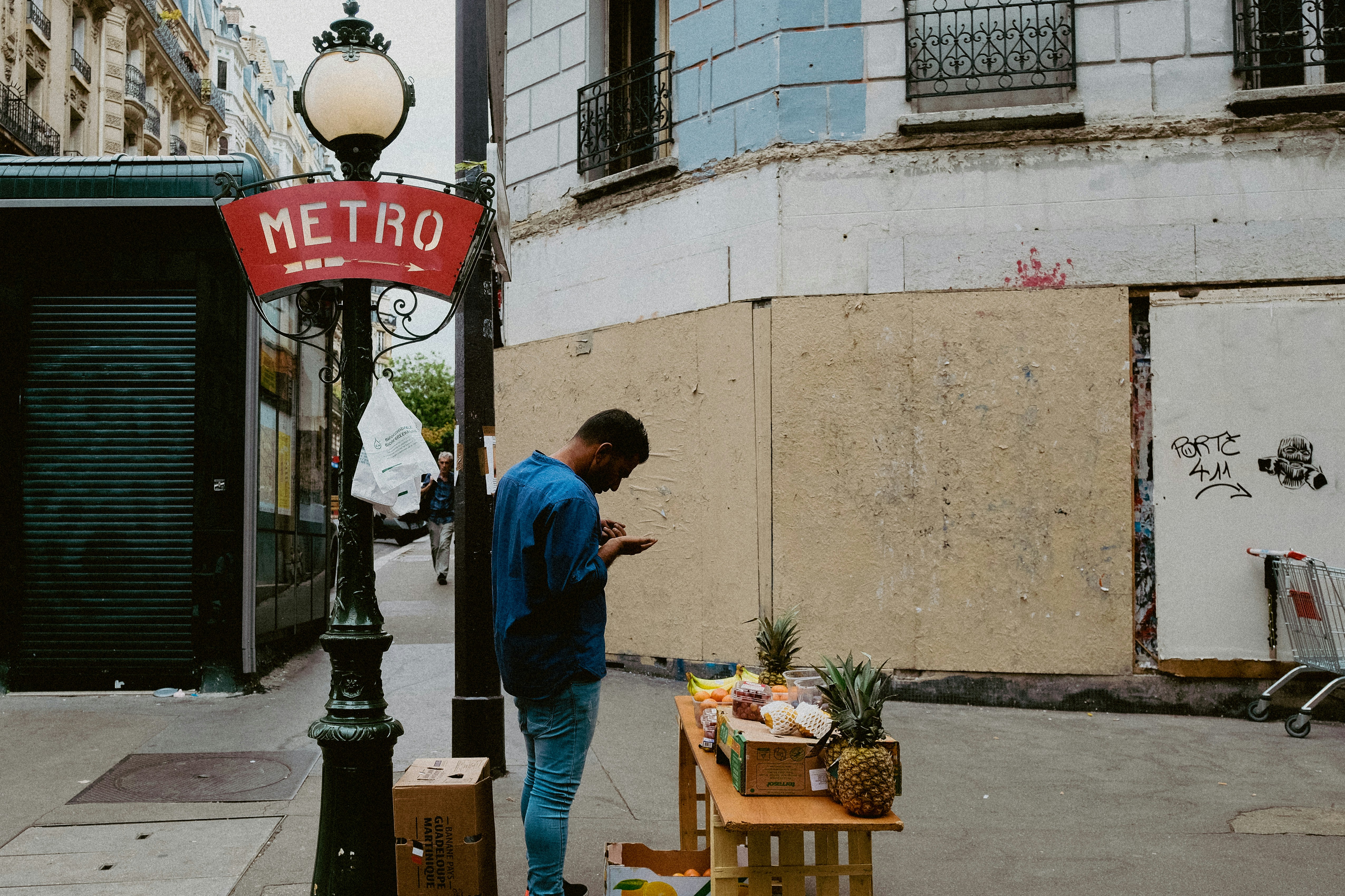 Man browsing phone beside a street fruit stall with pineapples and bananas under a Metro sign in a city setting.
