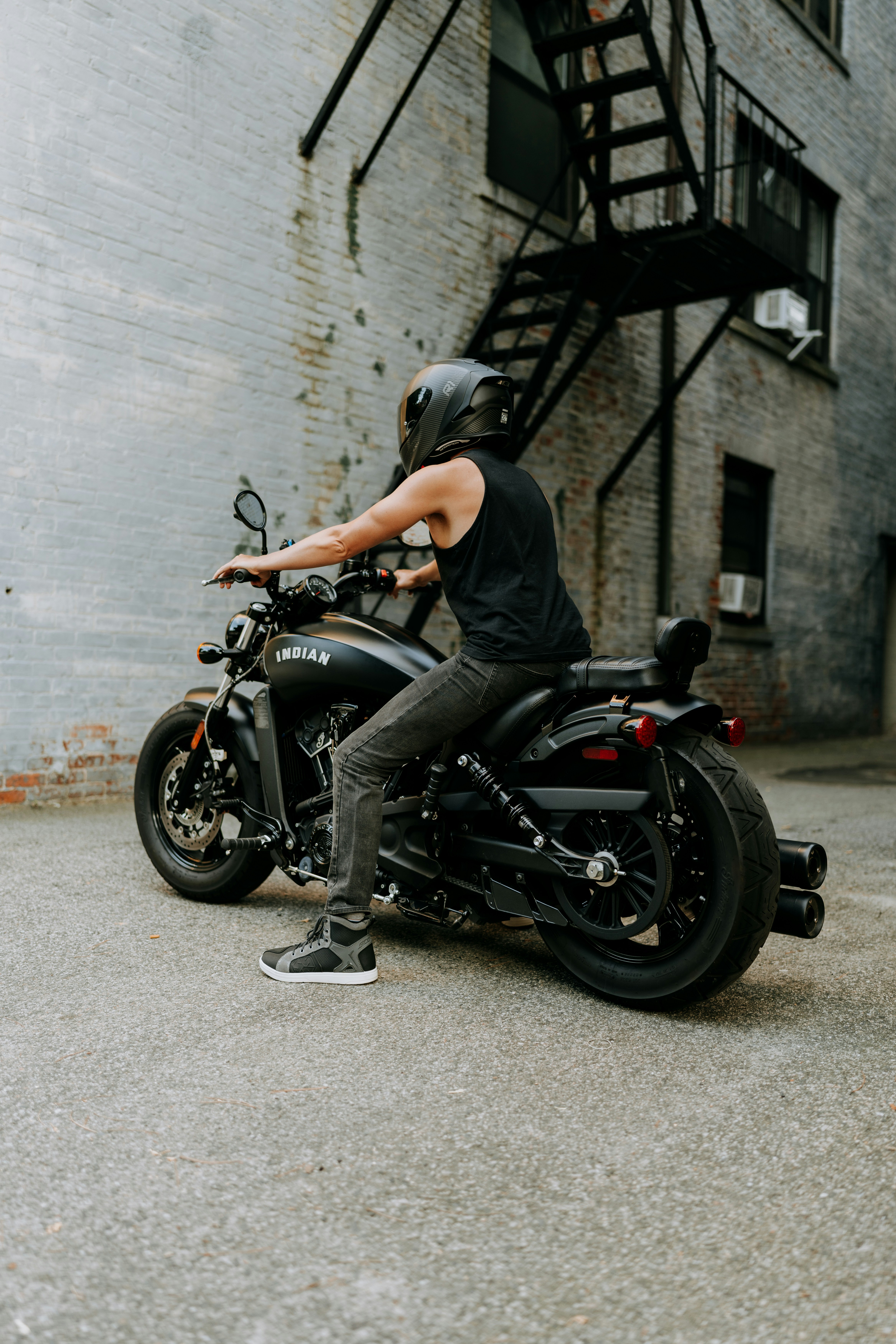 A motorcyclist poised on a sleek black Indian motorcycle, set against a textured brick wall in an urban environment.
