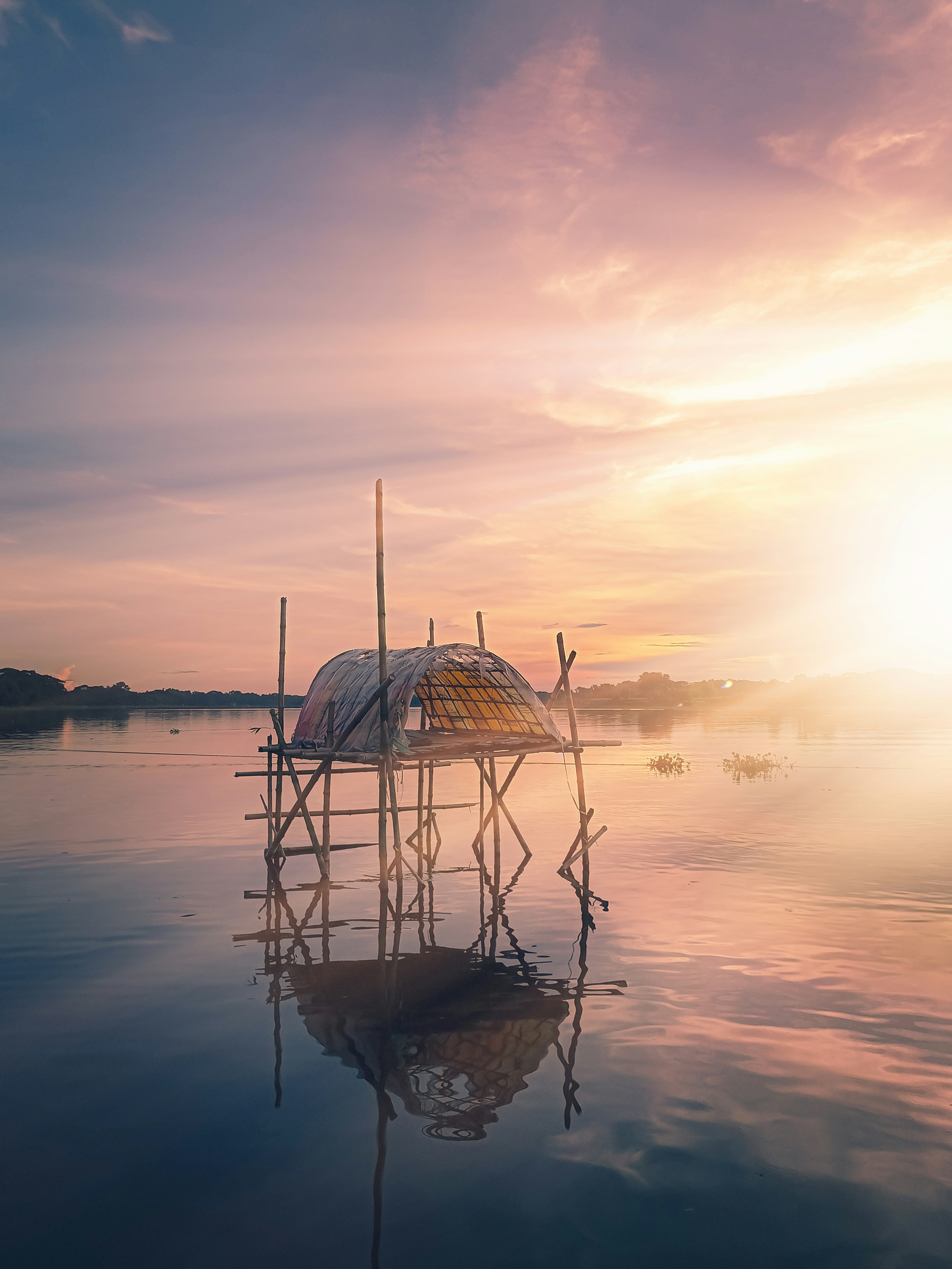 A wooden hut perched on stilts over calm waters, reflecting the vibrant hues of a sunset sky.