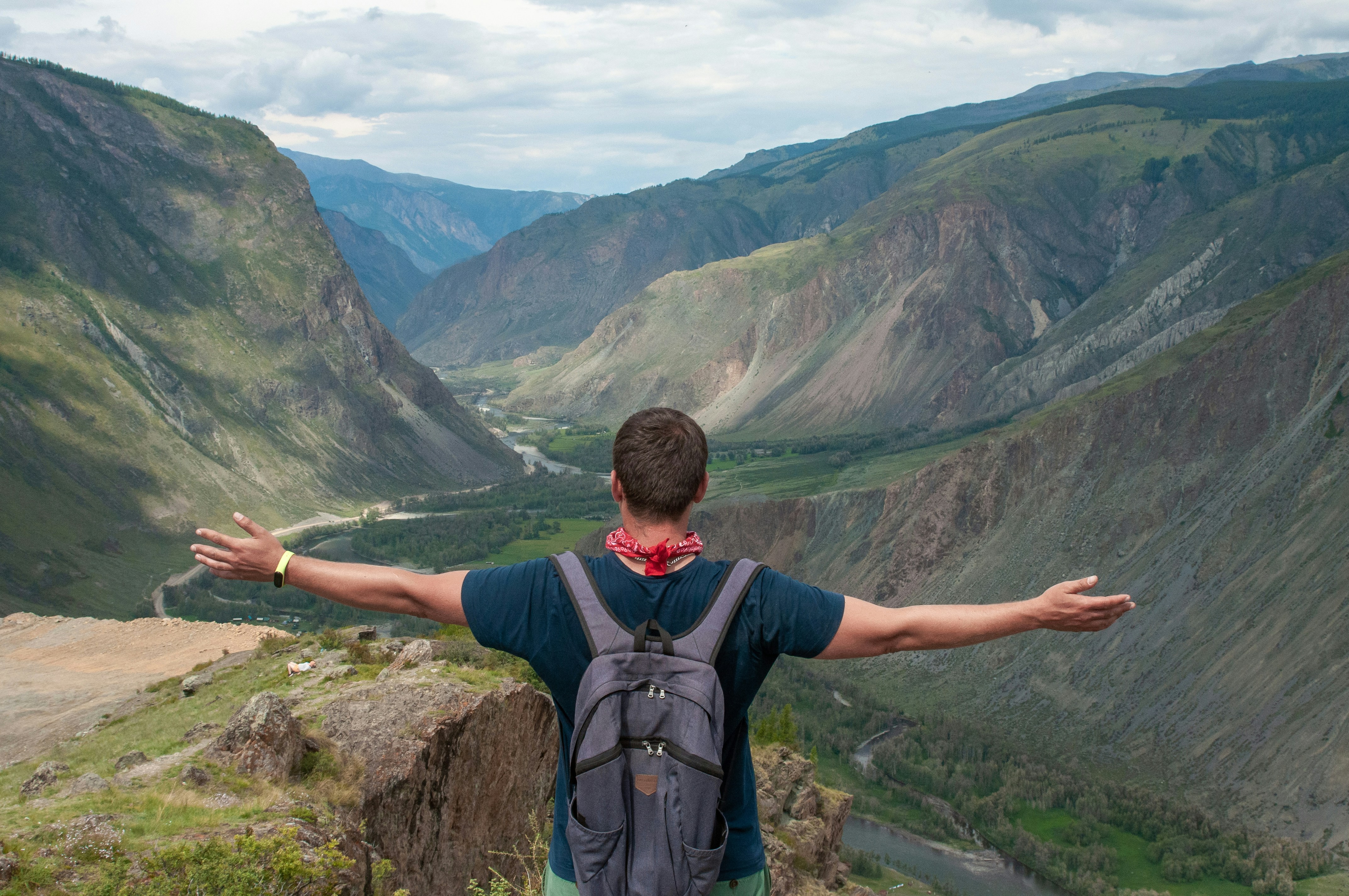 Person standing on a cliff with arms outstretched, overlooking a vast valley surrounded by mountains. The scene captures a moment of connection with nature.