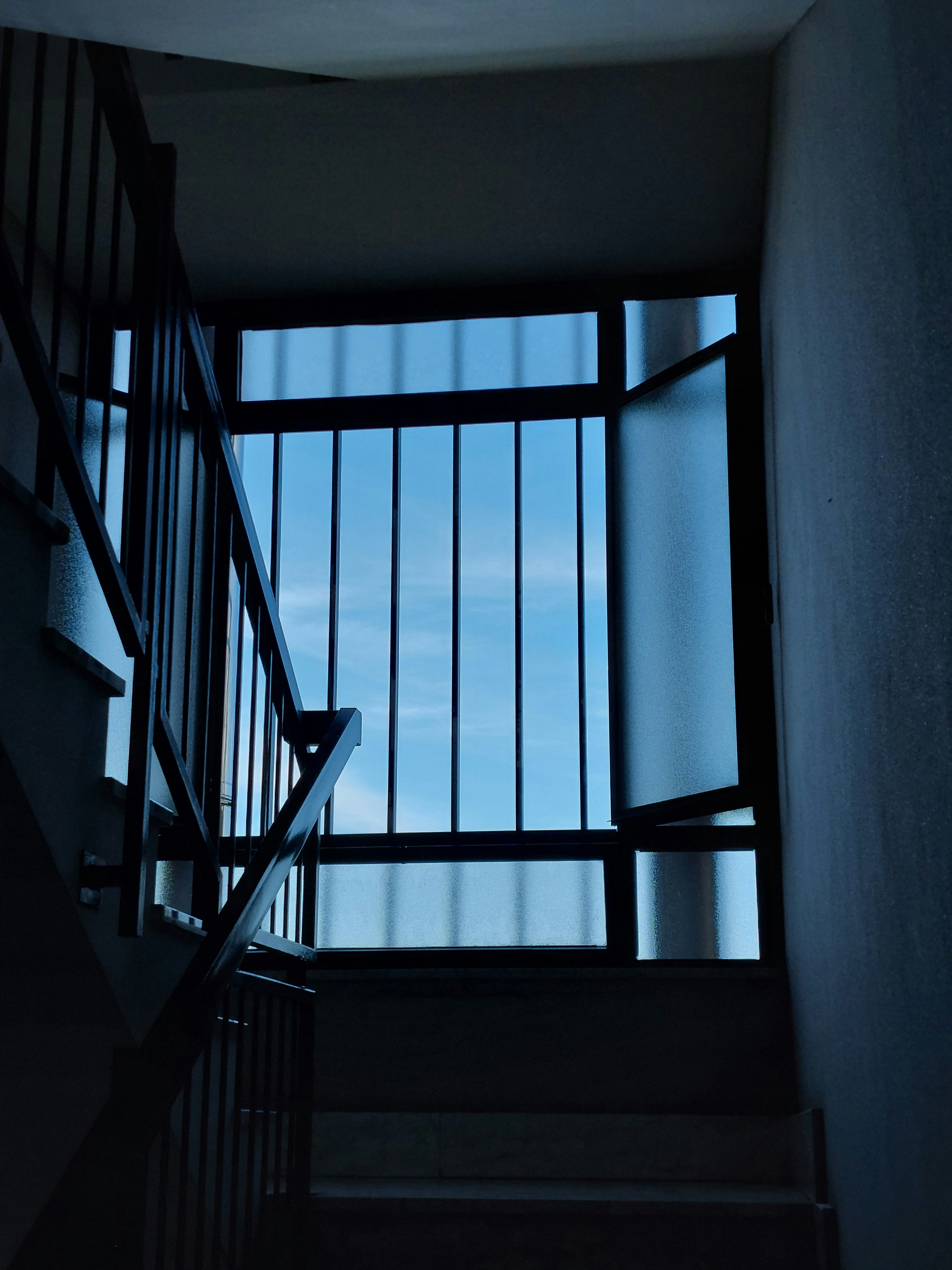 Staircase leading to an open window, framed by metal bars, with a clear blue sky visible beyond. The contrast between the interior shadows and the bright exterior creates a striking visual narrative.