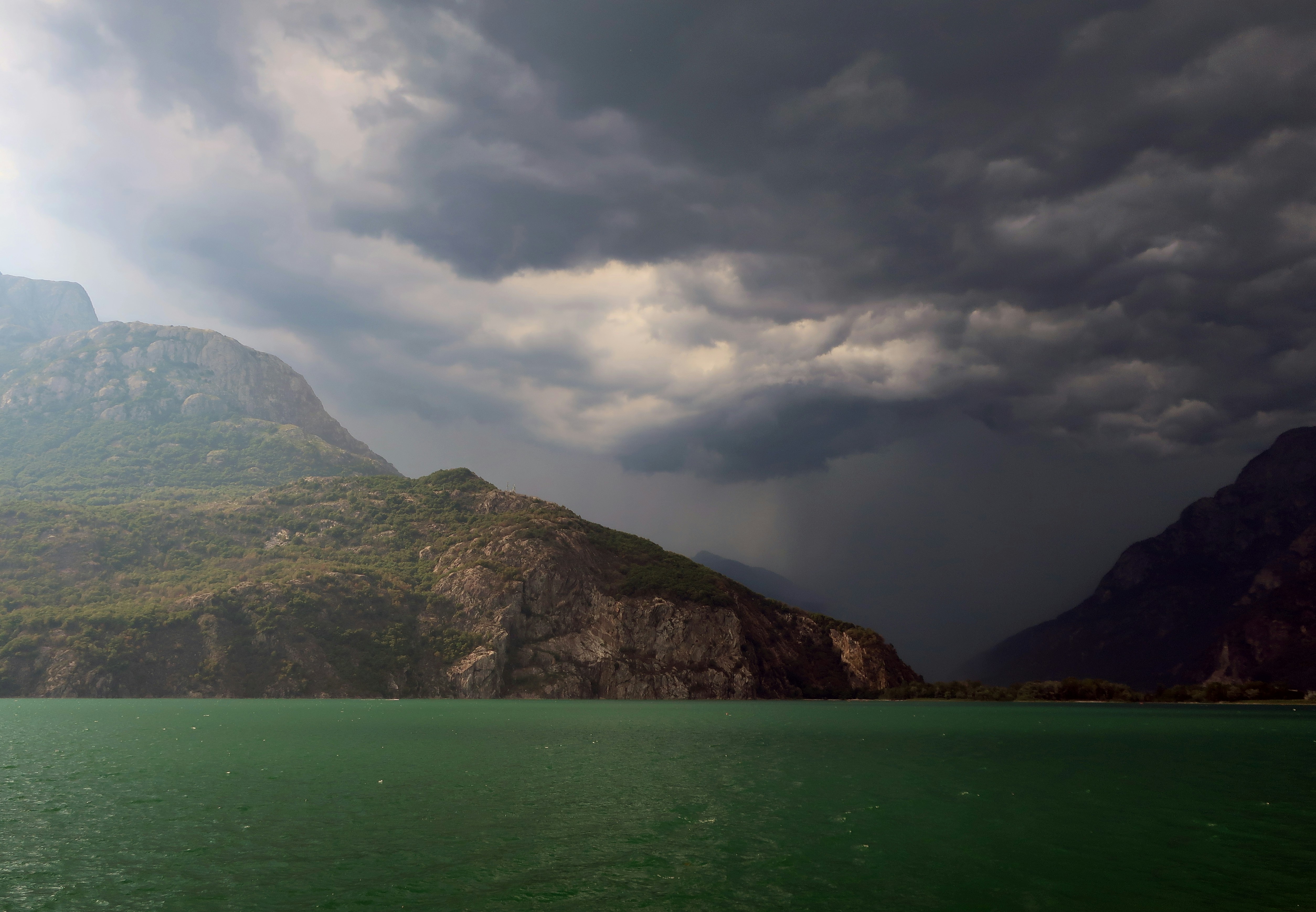 Ominous storm clouds gather over a rocky hillside by a green lake.