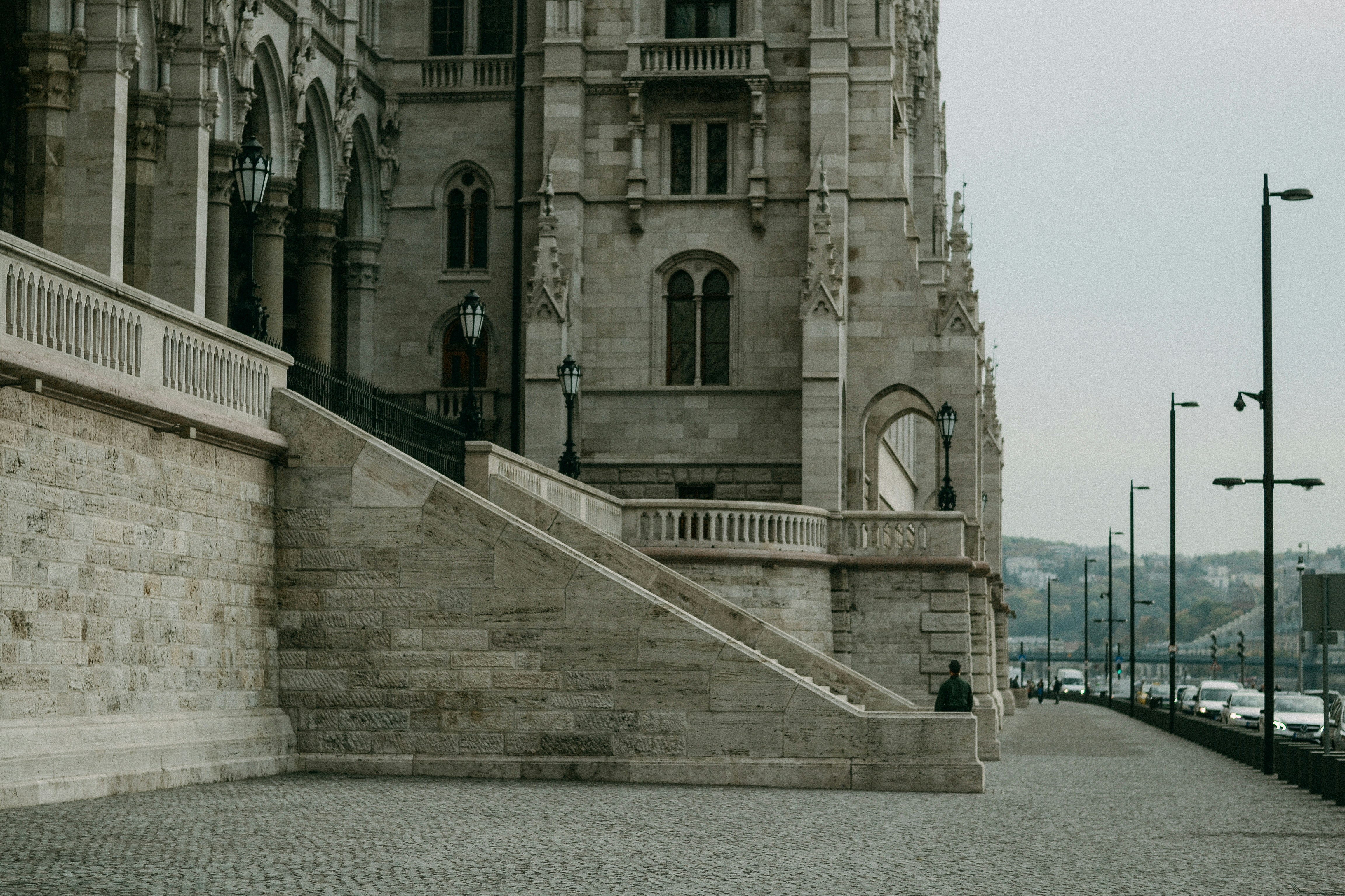 A solitary figure stands at the base of an imposing stone staircase, framed by intricate architectural details of a grand building. The scene captures a moment of quiet reflection amidst urban architecture.
