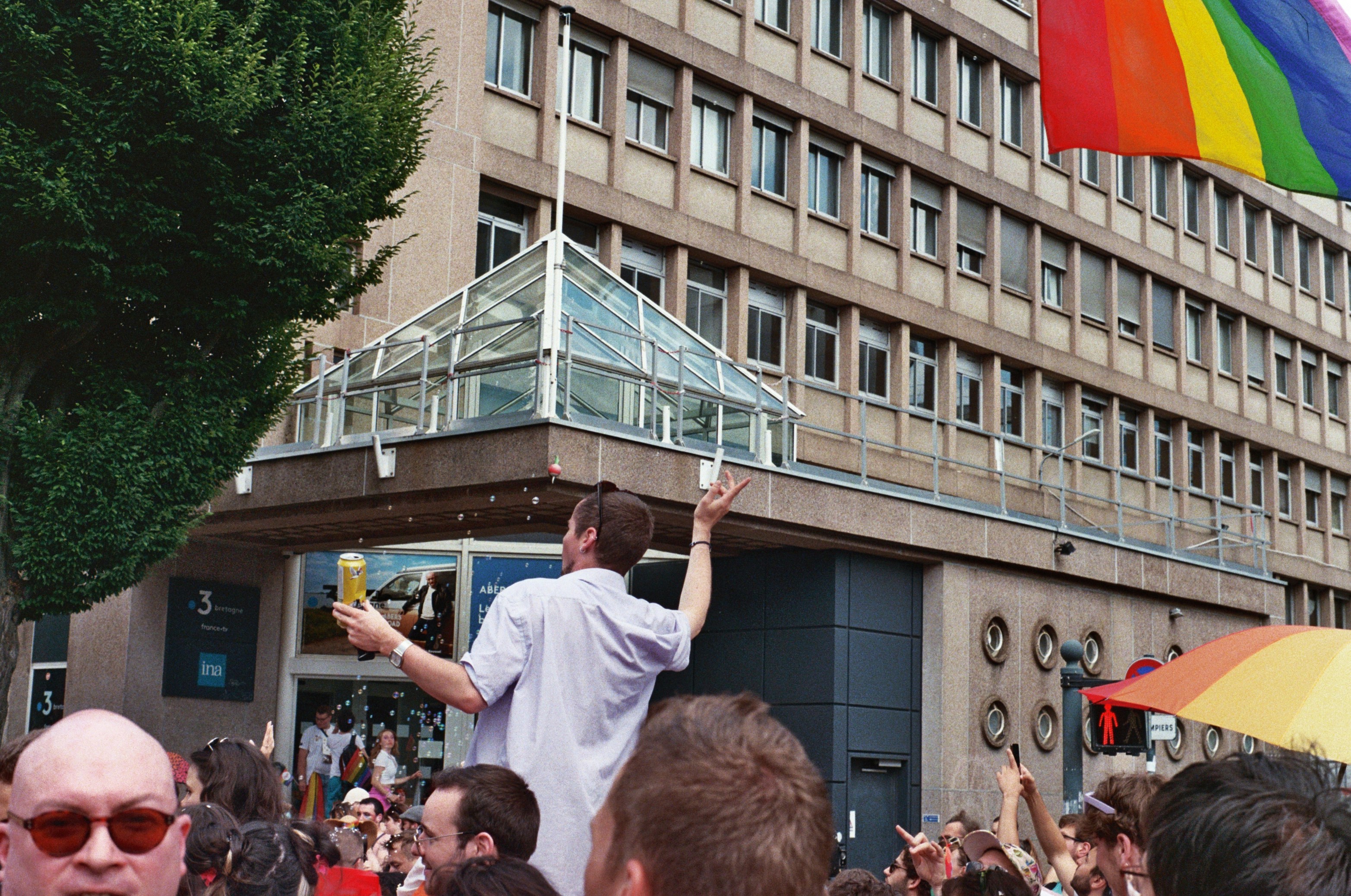 Crowd gathered outside a building during a festive event, with a person raising a drink and a rainbow flag waving in the background.