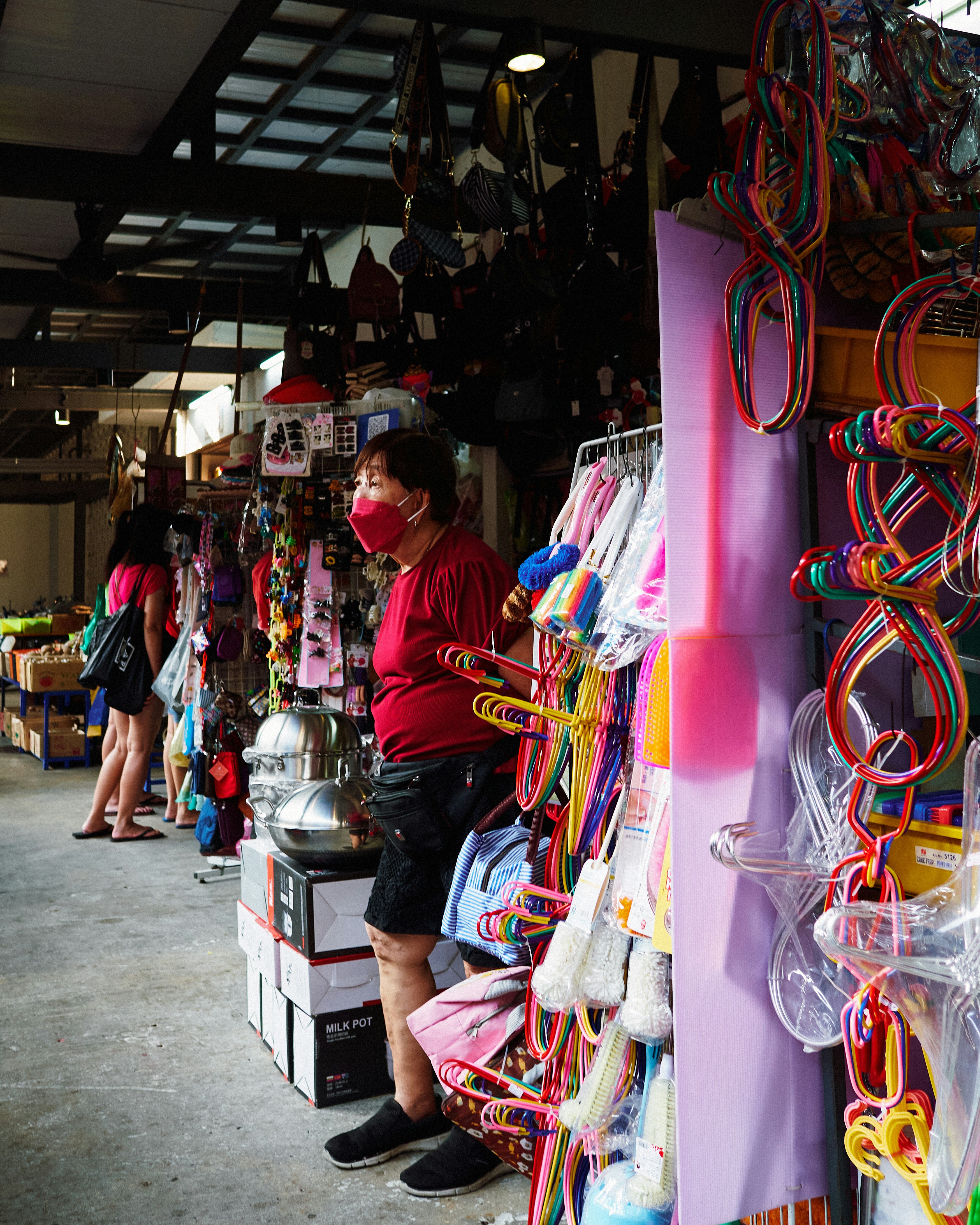 A bustling market scene featuring a vendor in a red shirt, surrounded by colorful merchandise and shoppers. The atmosphere is lively and engaging.