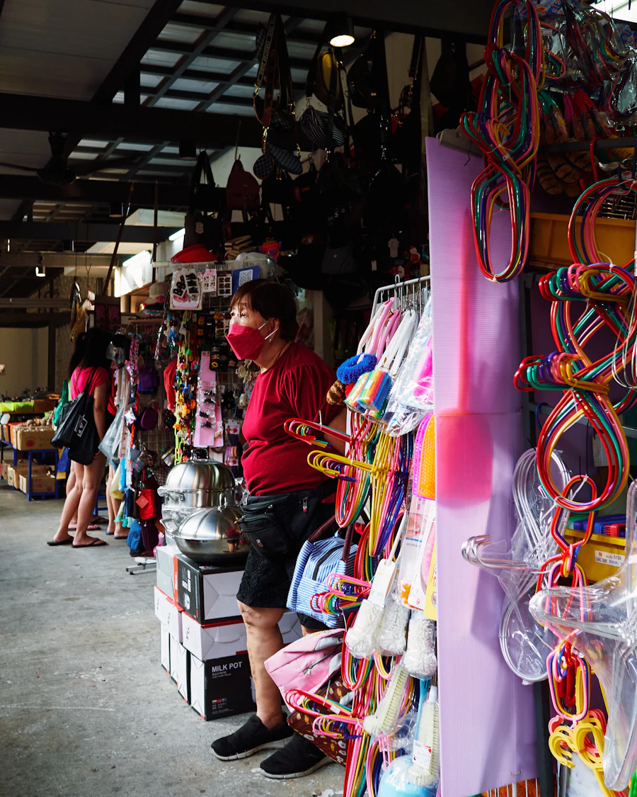 A masked vendor stands beside a shop packed with bright plastic hangers and household goods. Another shopper browses in the background under a dim market roof.
