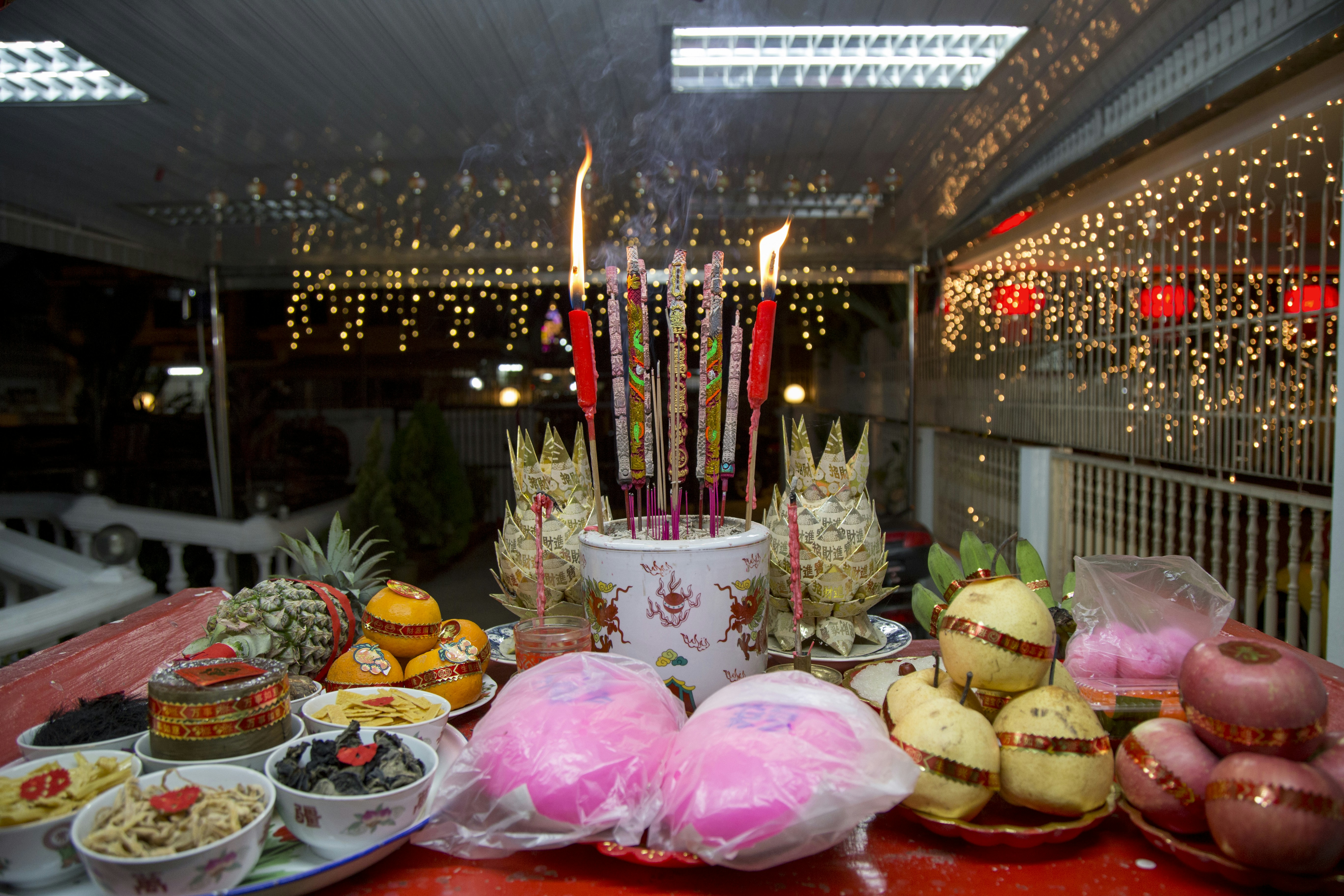 An array of colorful offerings, including fruits, sweets, and incense, arranged on a table for a festive celebration. The background features twinkling lights, enhancing the festive atmosphere.