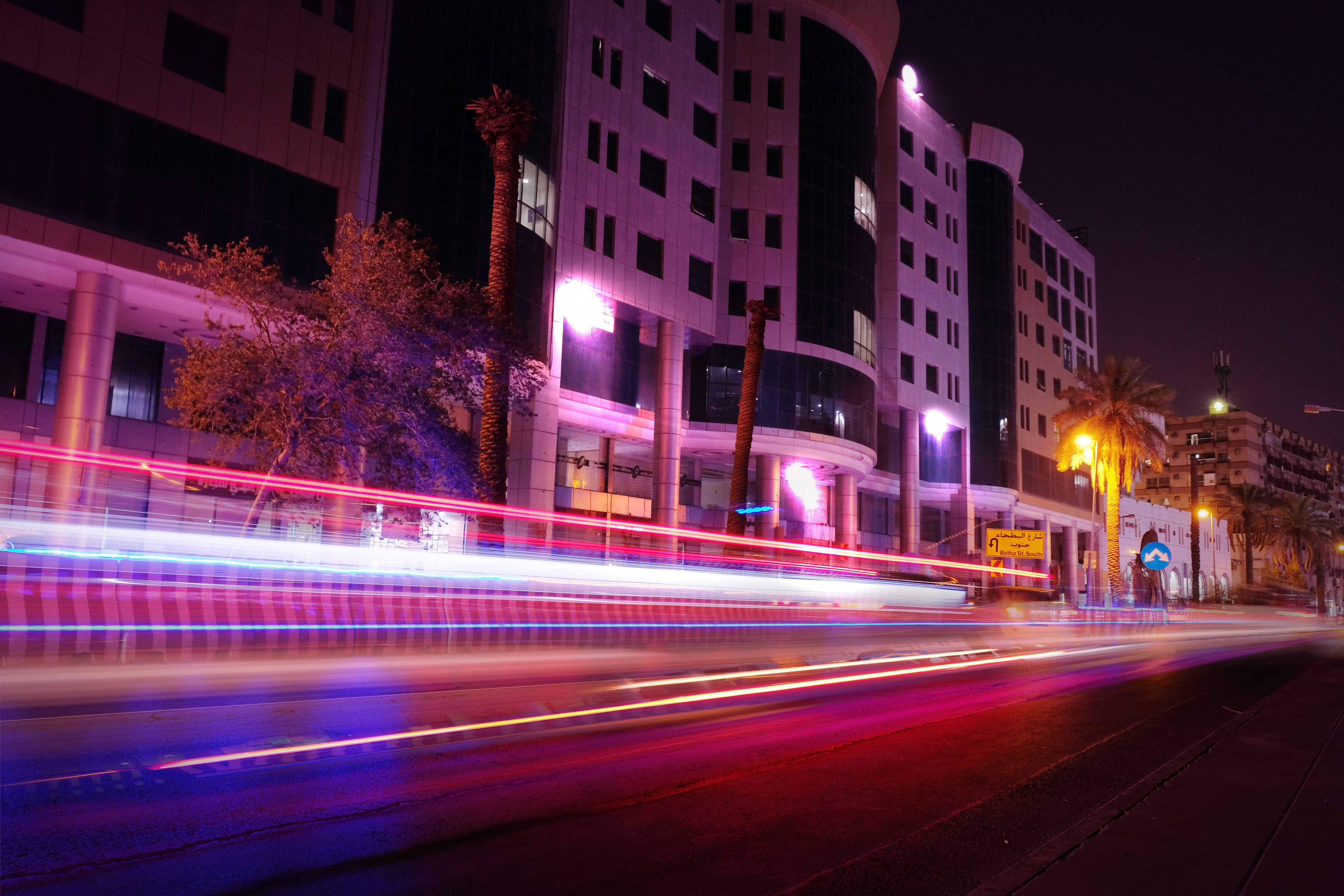 Nighttime street scene with light trails from passing vehicles against illuminated buildings and palm trees.