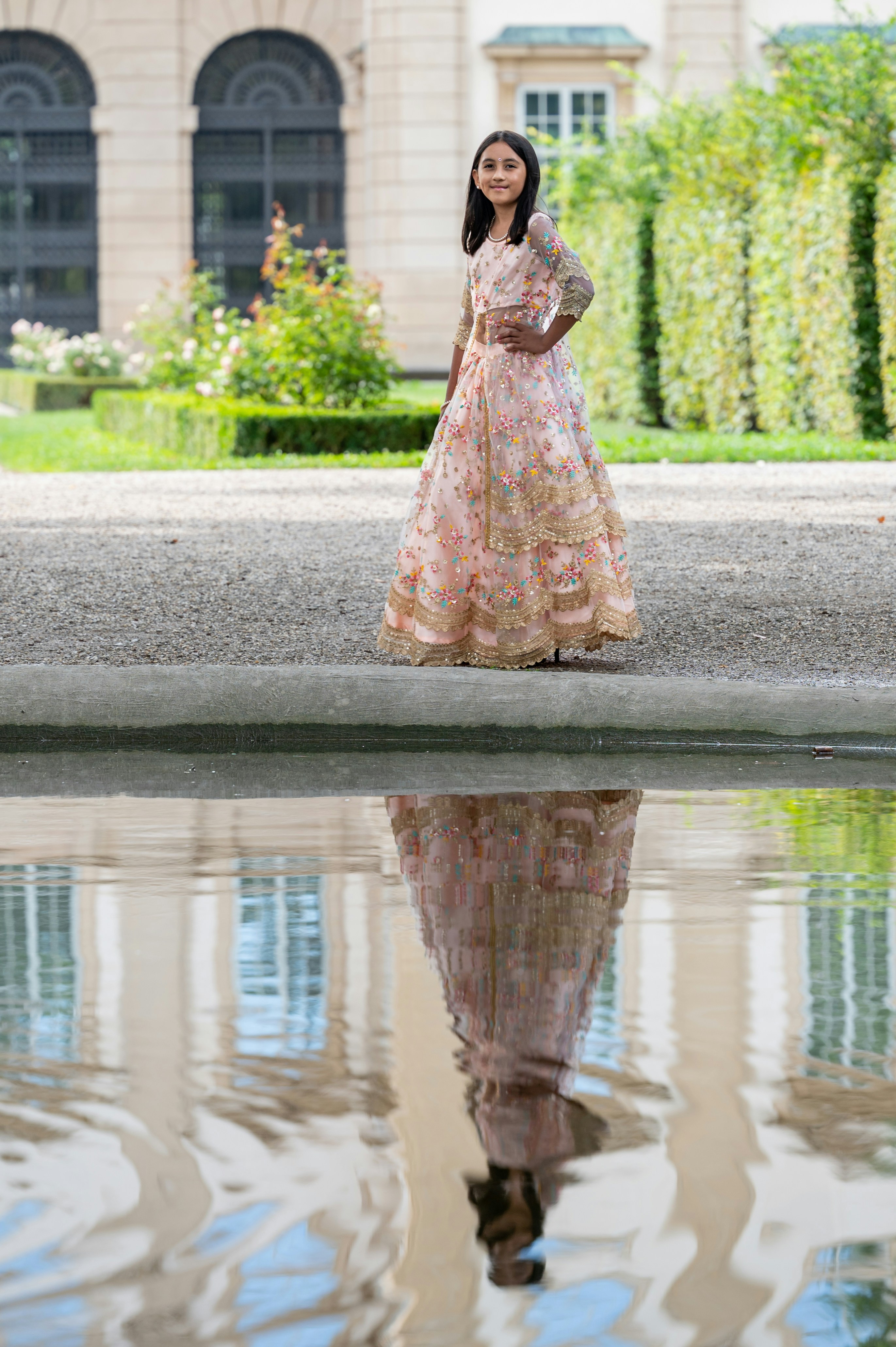 Young girl in a beautifully embroidered pink dress poses gracefully by a reflective water feature in a lush garden setting.