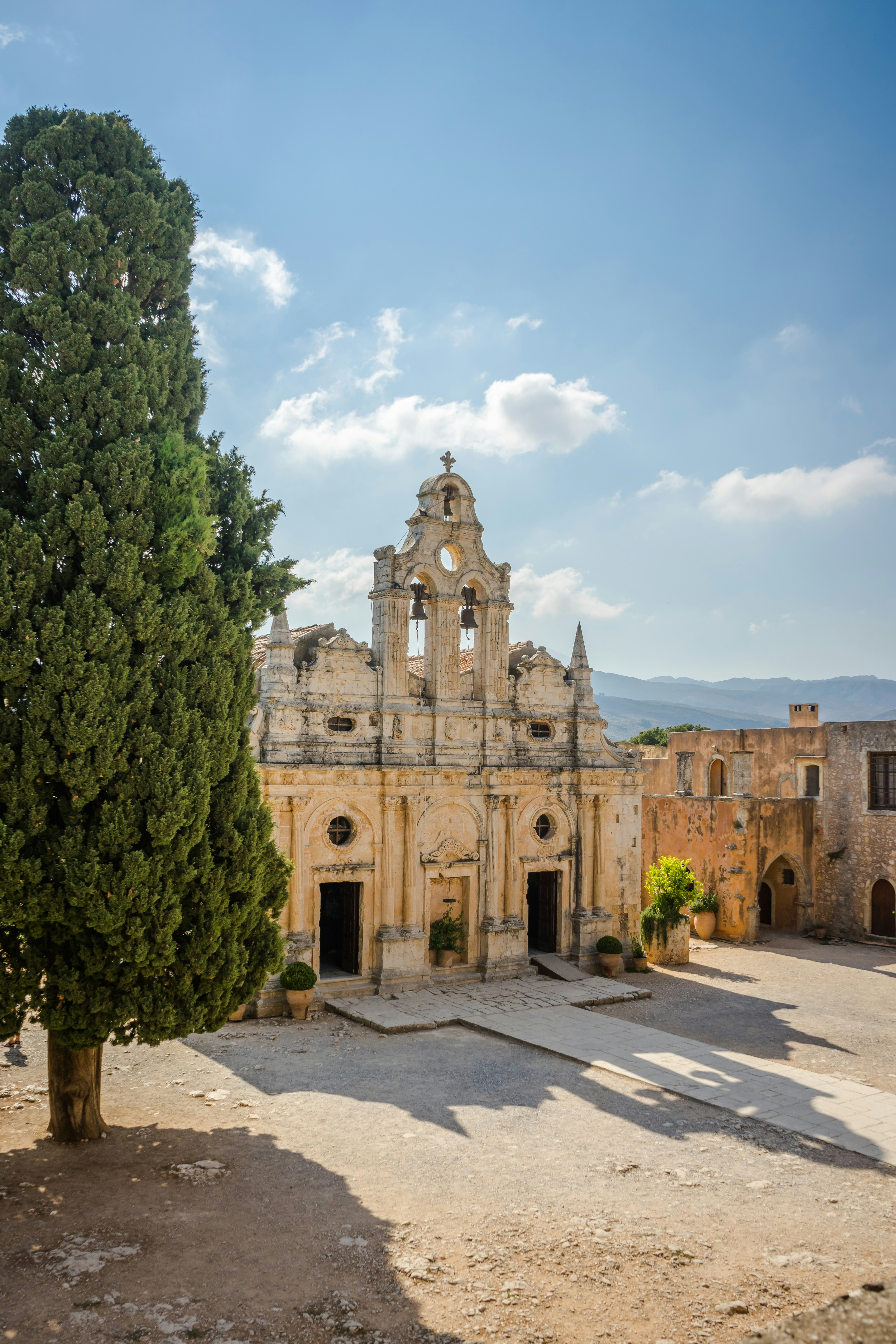 Imposing monastery structure framed by a lush cypress tree, set against a clear sky. The scene captures the serene beauty of ancient architecture in a tranquil courtyard.
