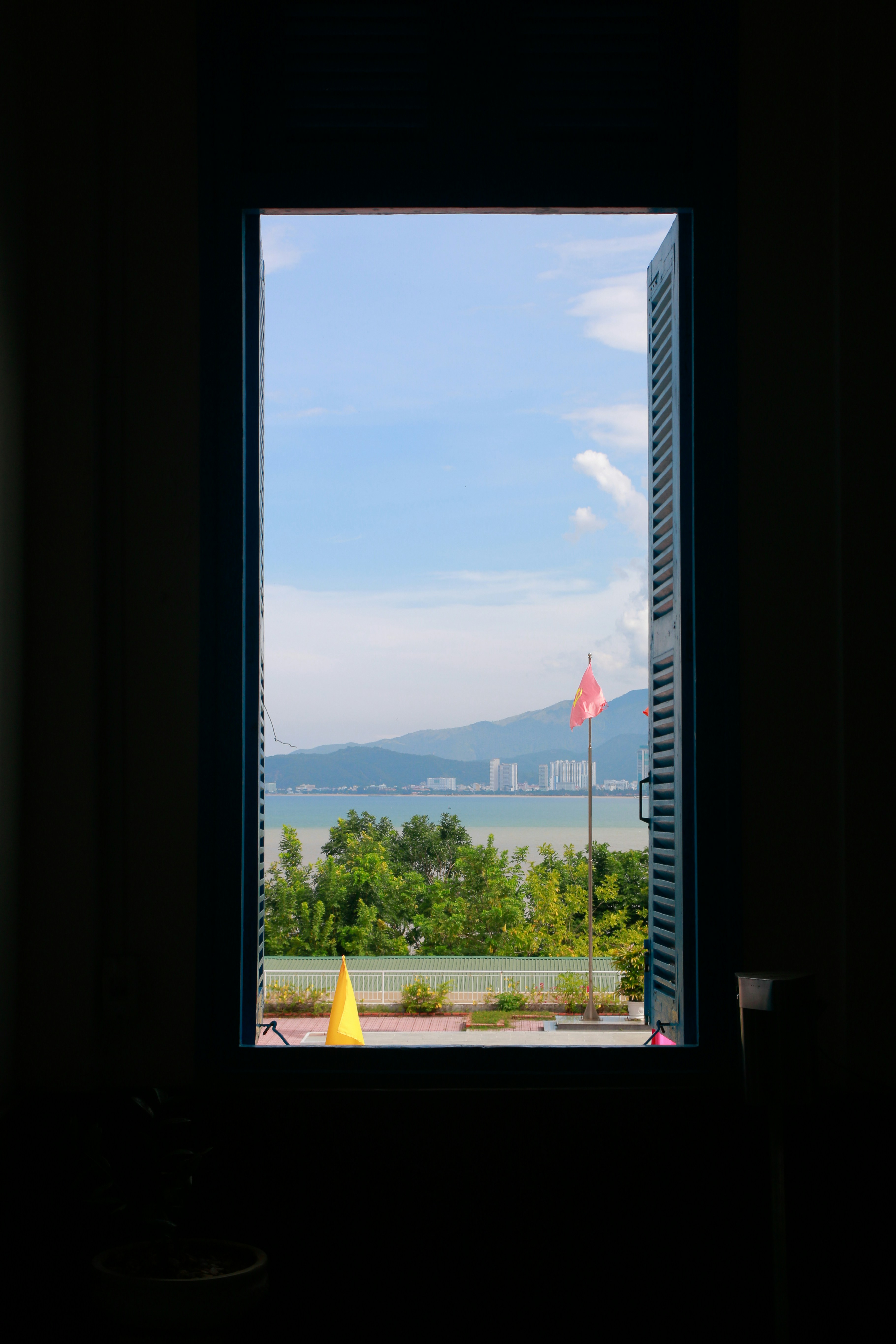 Open shutters reveal a tranquil coastal scene, featuring lush greenery and distant mountains under a clear sky. A flag flutters gently in the breeze.