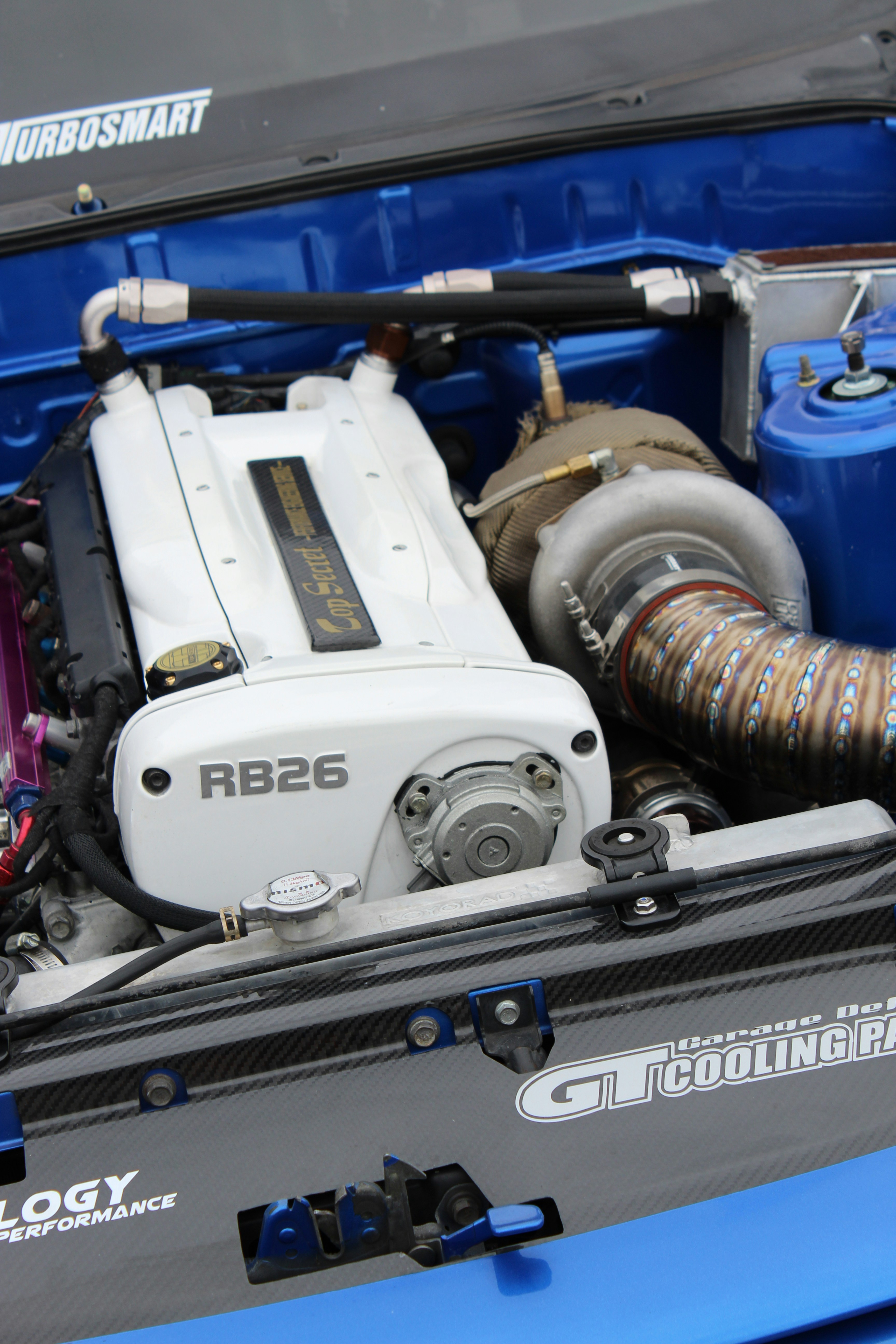 Close-up photograph of a Nissan RB26 turbocharged engine under a vivid blue hood, highlighting the large turbo and polished intake. The scene showcases carbon-fiber panels and performance-focused hardware.