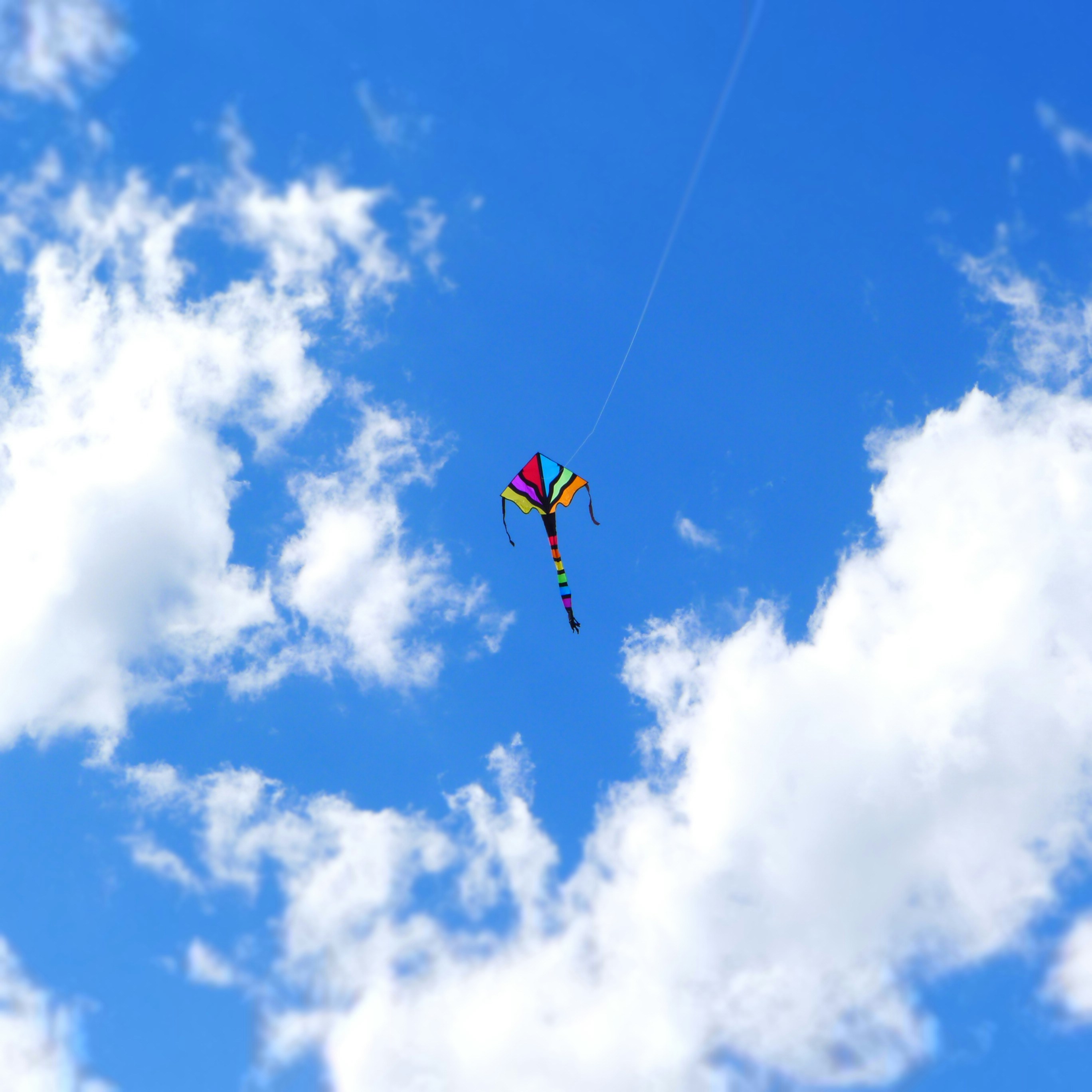 Colorful kite soaring high amidst fluffy clouds in a bright blue sky.