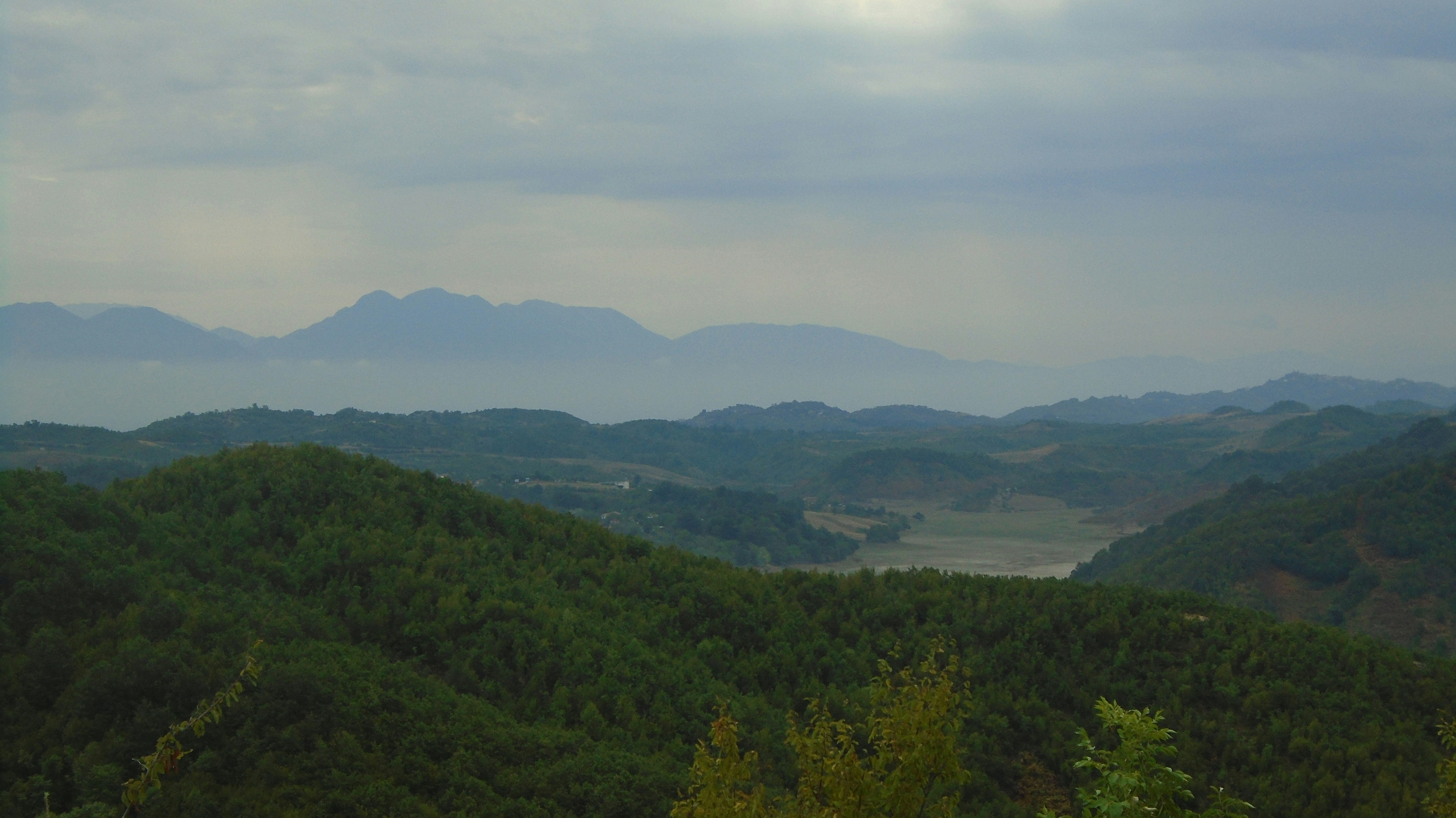 Vast rolling hills under a cloudy sky, with distant mountains fading into the mist. The landscape showcases the serene beauty of nature's undulating terrain.