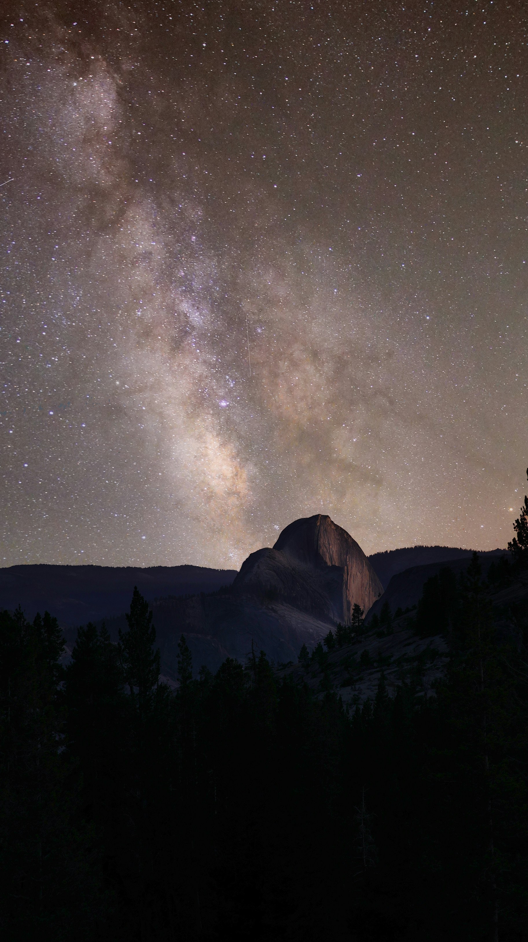 Milky Way galaxy illuminating the night sky above a prominent granite peak, framed by dark silhouettes of surrounding trees.