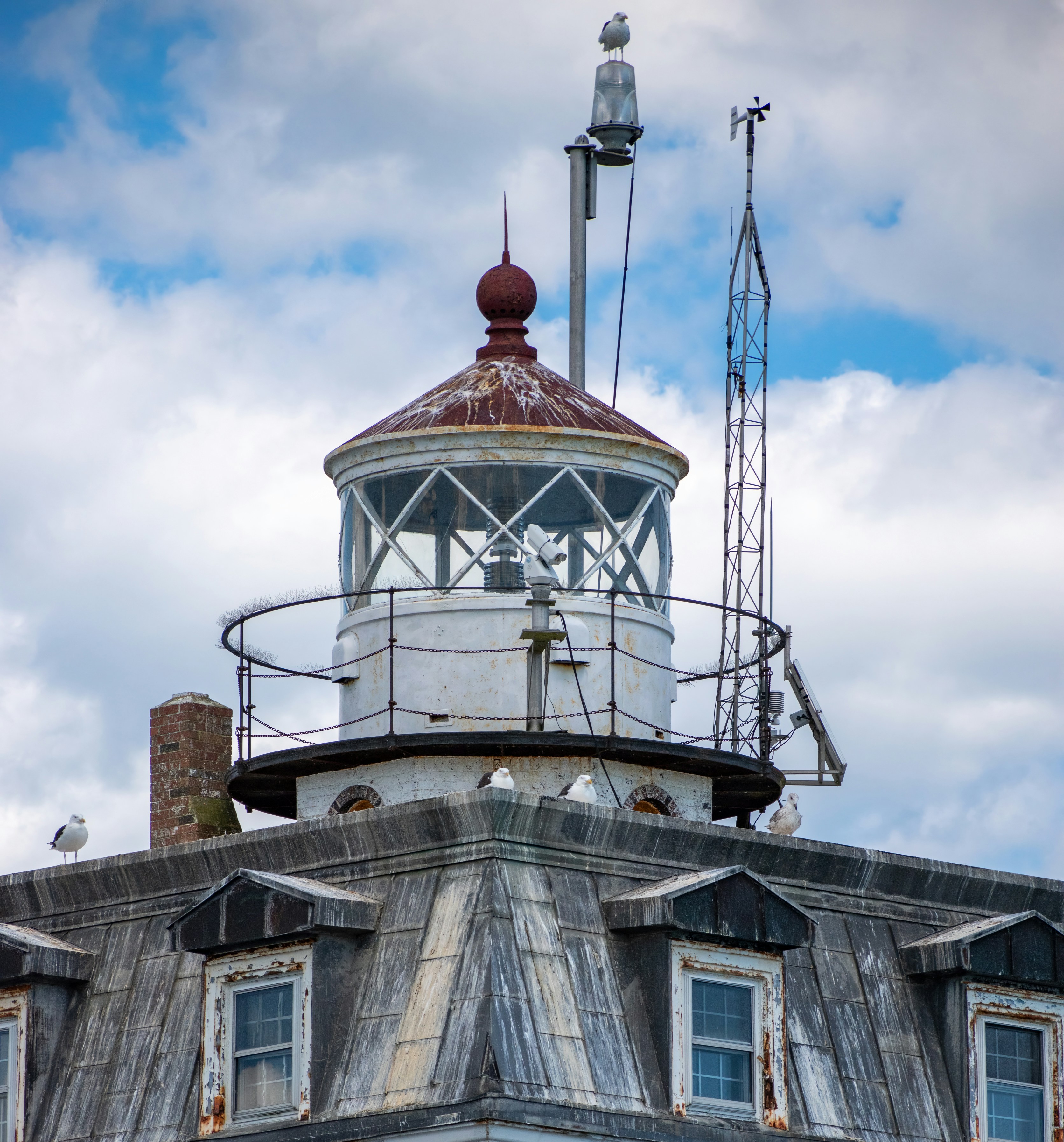 Weathered lighthouse with a distinctive red dome and surrounding antennas, set against a backdrop of cloudy skies.