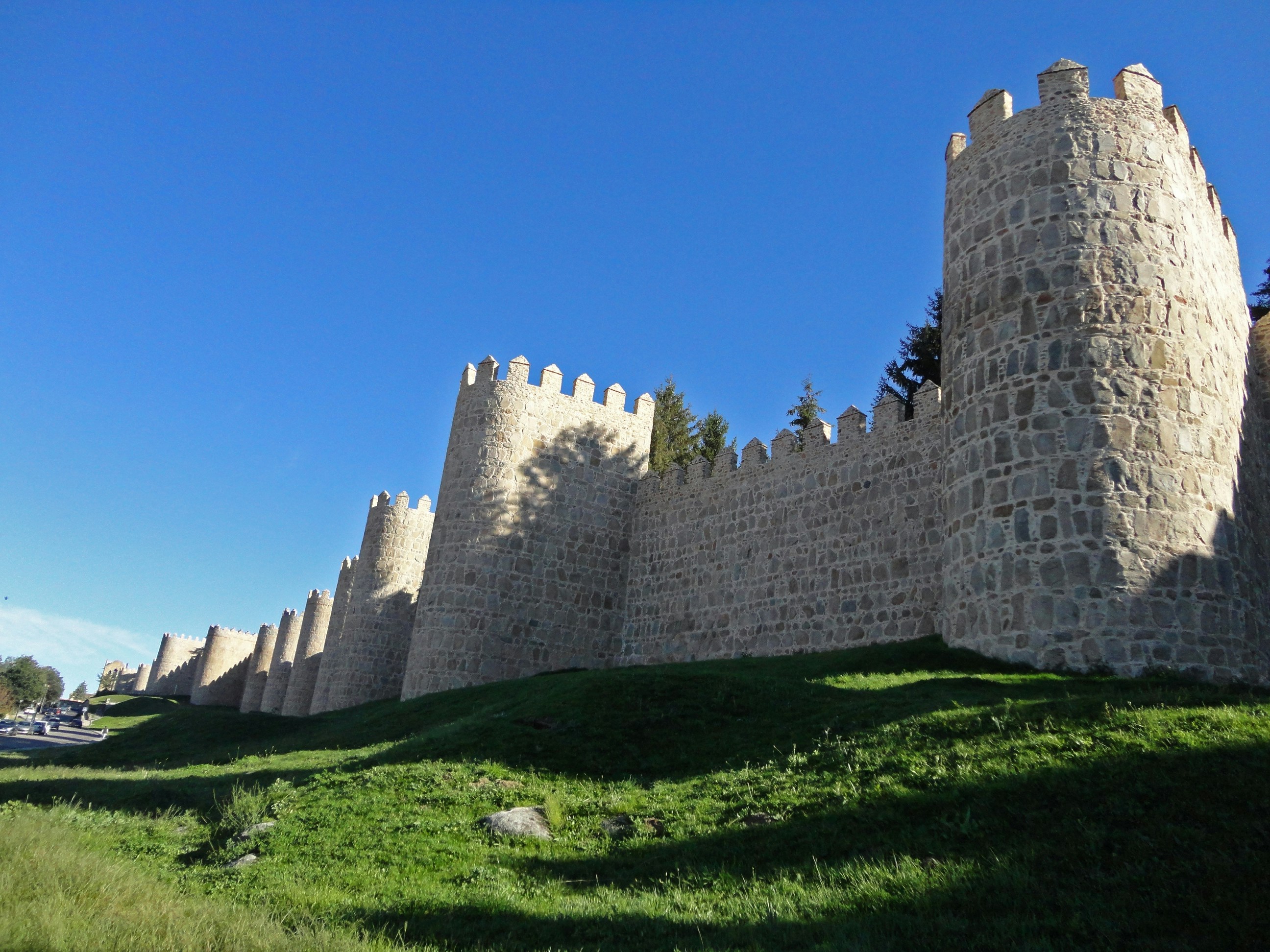 Historic stone ramparts and a round tower extend along a sunlit grassy slope beneath a clear blue sky.
