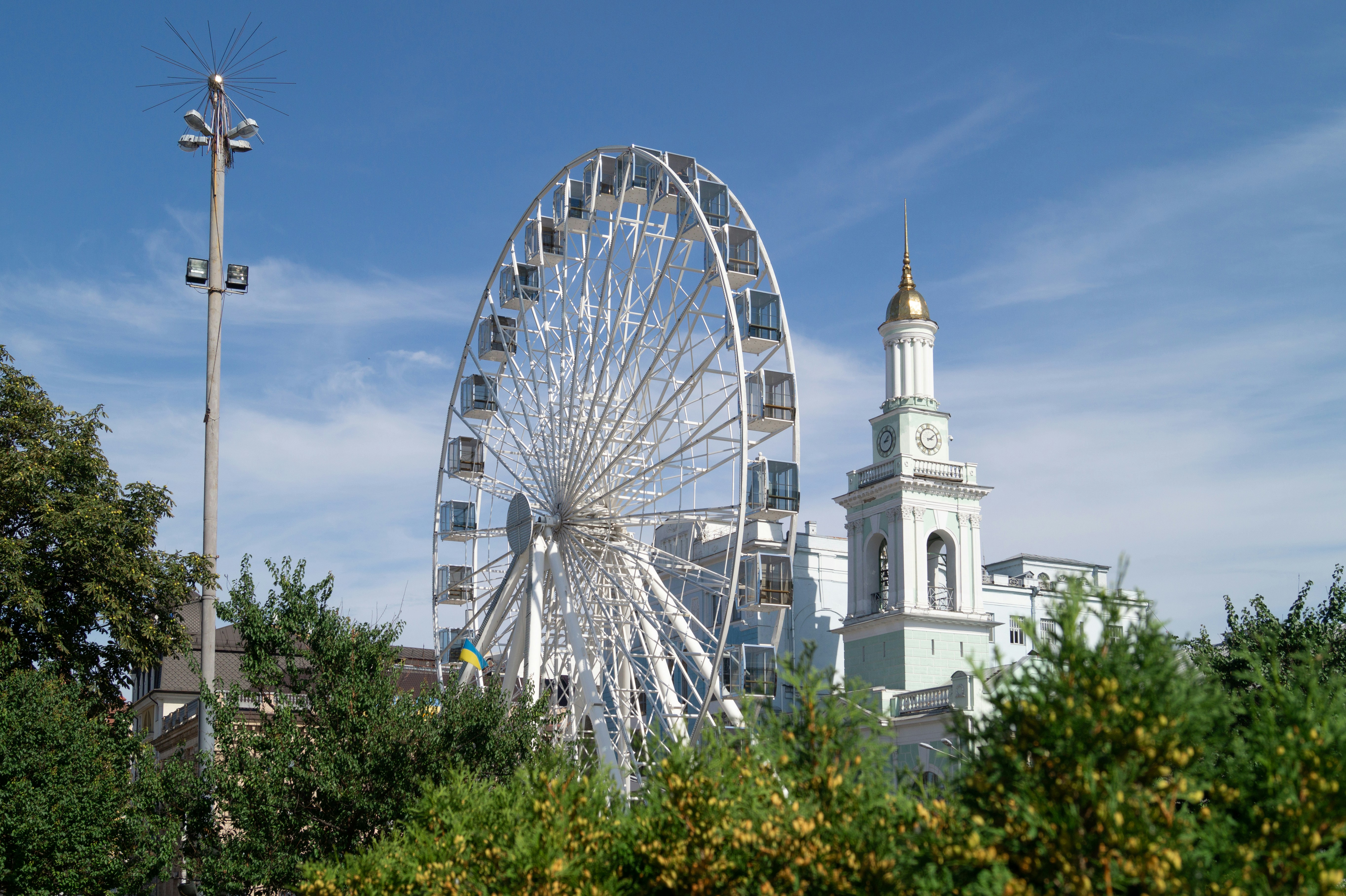A towering Ferris wheel stands beside a historic clock tower, framed by lush greenery under a clear blue sky.