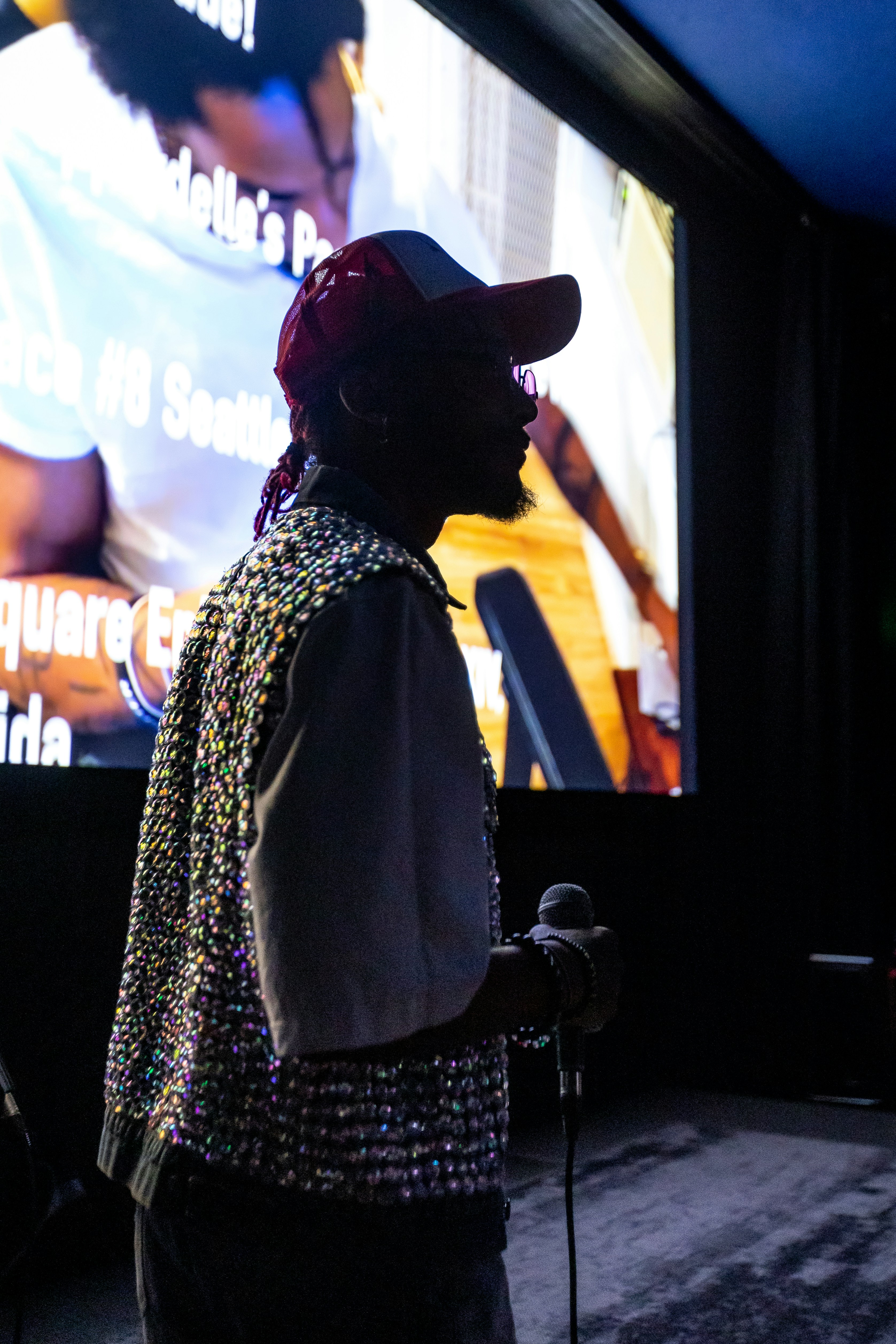 A performer in a sequined vest stands on stage, microphone in hand, against a backdrop displaying vibrant visuals. The atmosphere is charged with creativity.