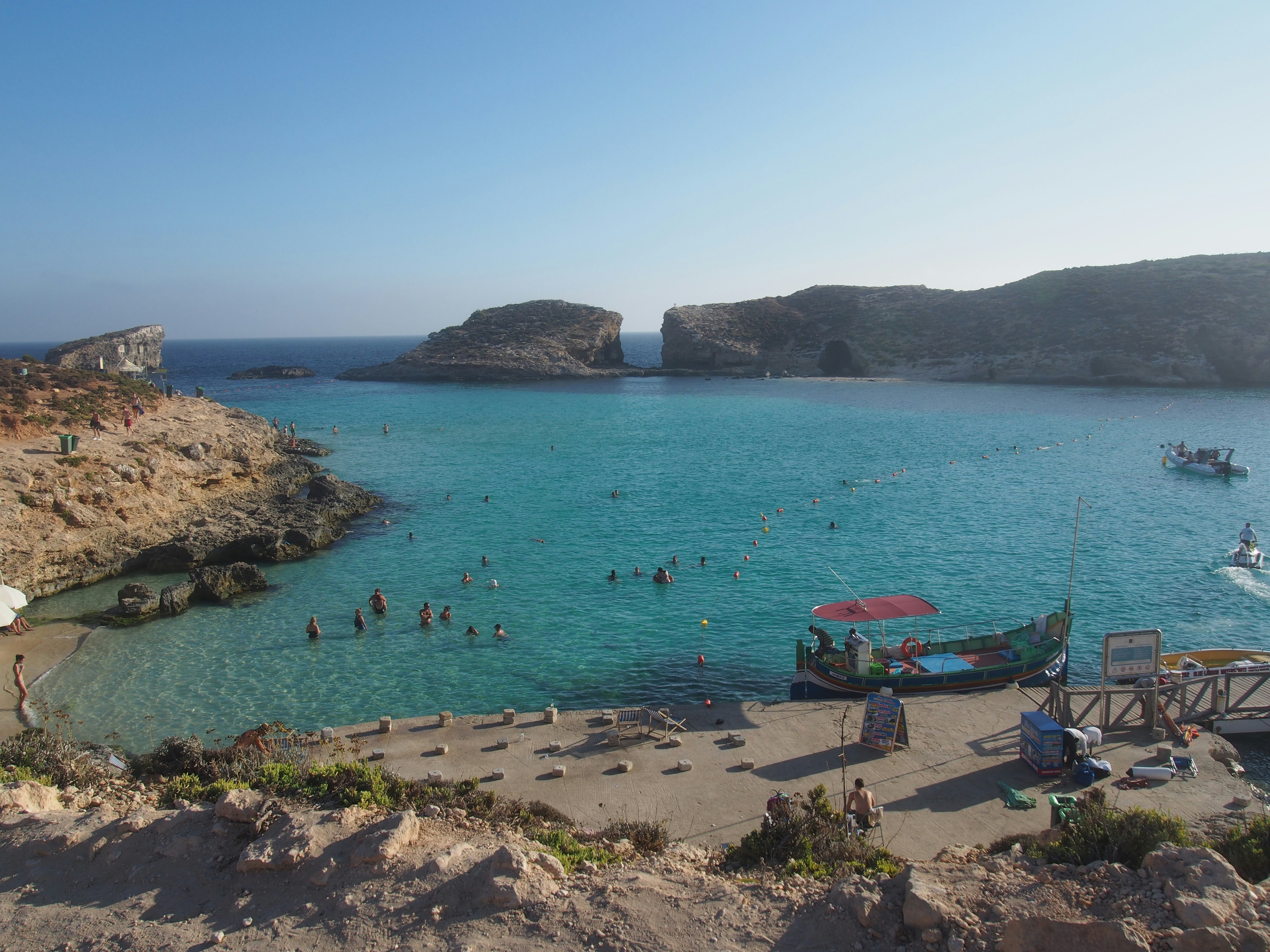 Turquoise cove crowded with swimmers along a rocky shore, with a small dock and a red-canopy boat. Distant limestone arches frame the horizon.
