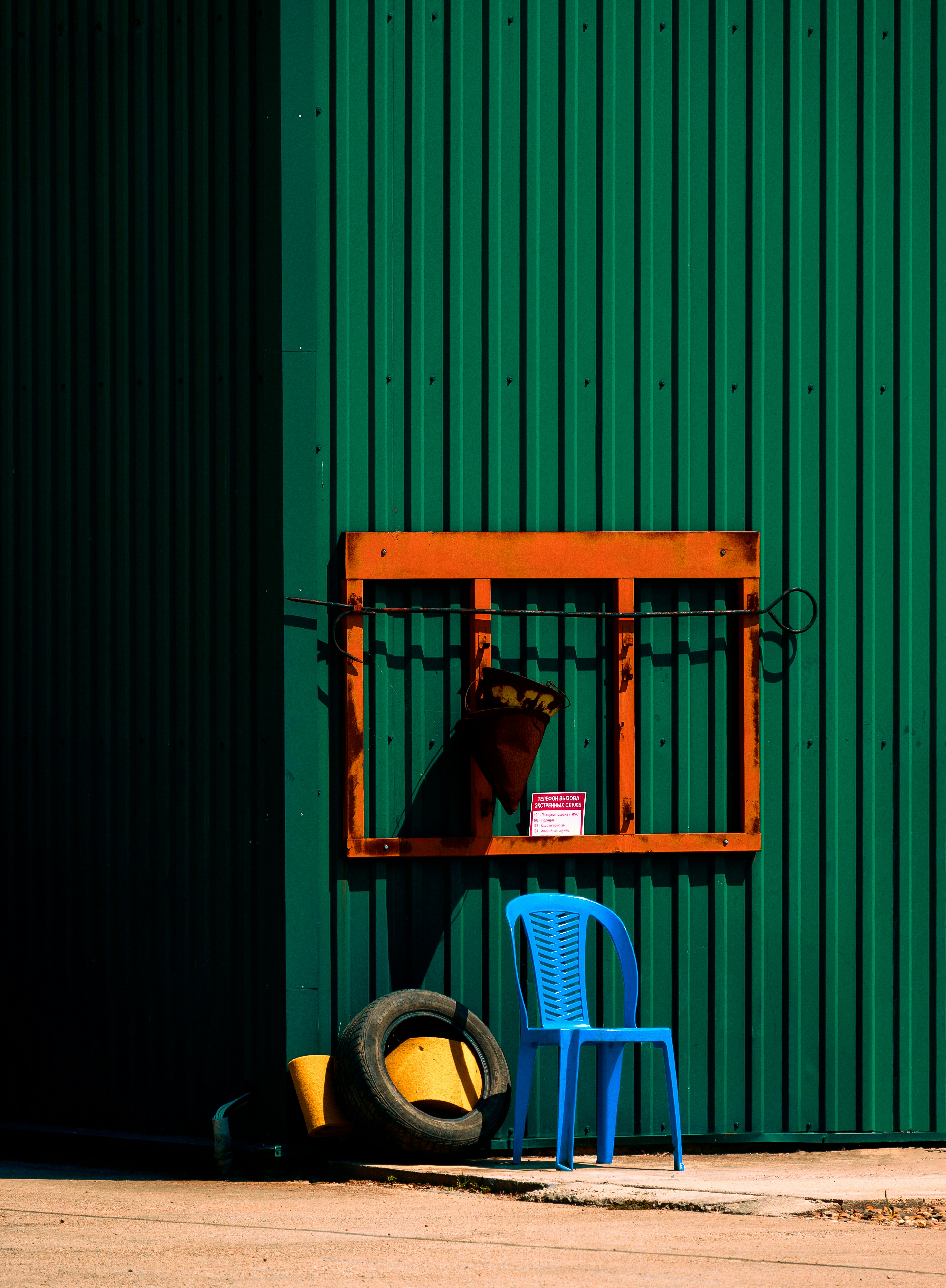 A vibrant blue plastic chair rests against a green corrugated wall, accompanied by a stack of tires and an orange window frame. A sign hangs nearby, adding to the scene's urban character.