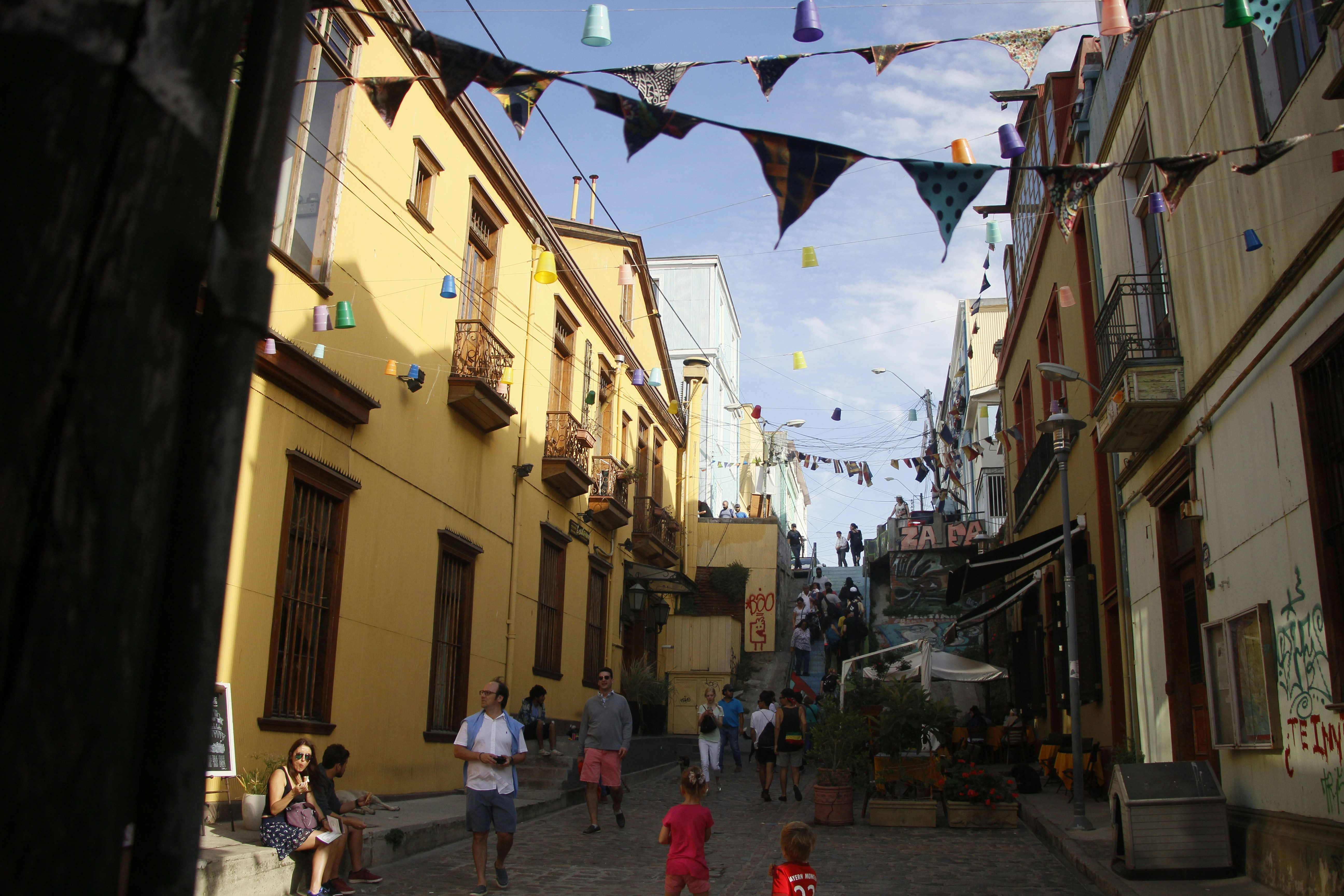 Colorful flags and bustling pedestrians in a narrow alley with yellow buildings.