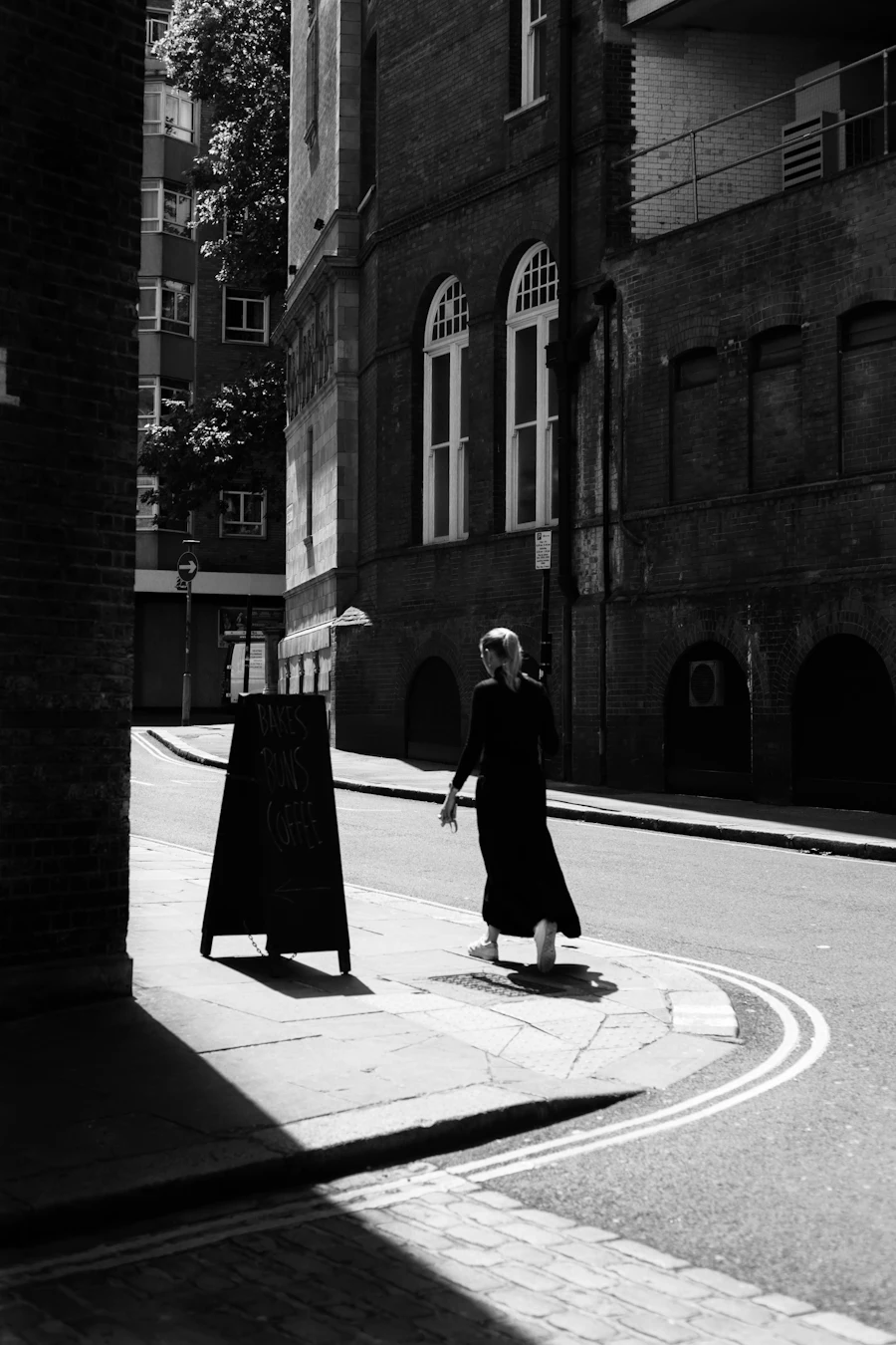 A lone woman in a long dark dress walks through a narrow city street in strong black-and-white contrast. Tall brick buildings and a sandwich board frame the sunlit sidewalk.