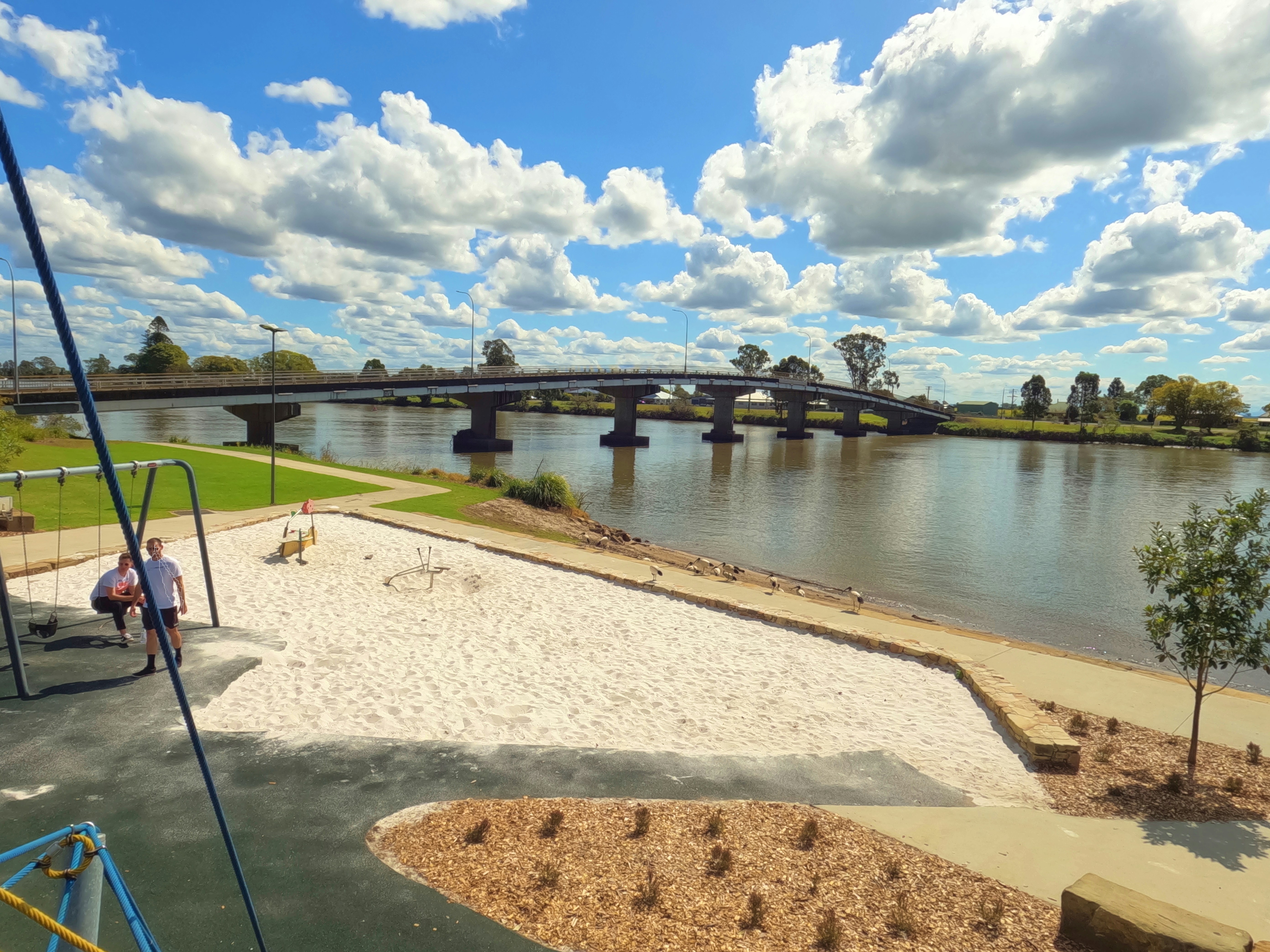 Bridge spanning a calm river with a sandy path and lush greenery beneath a sky dotted with clouds.