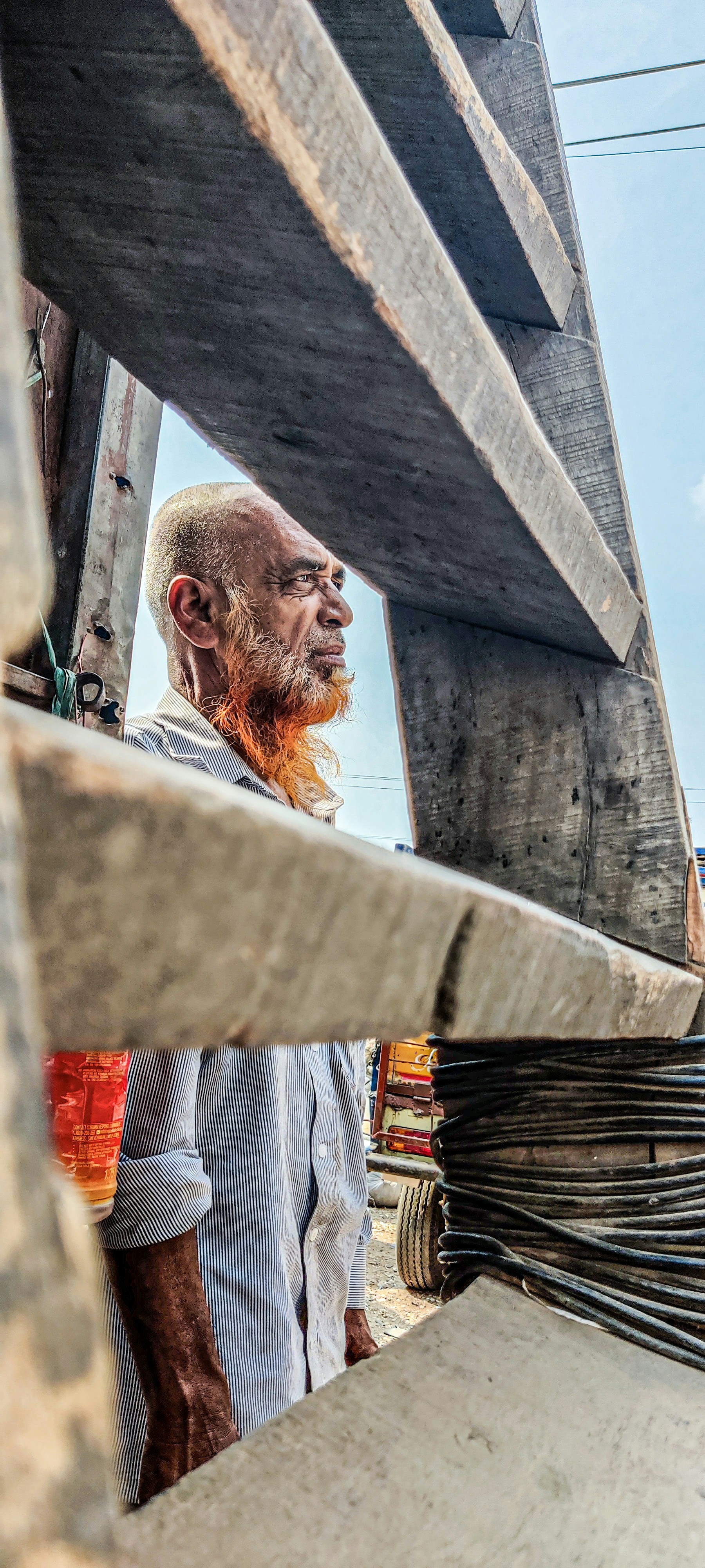 Portrait of an elderly man with an orange beard, framed by rough wooden beams at a construction site.