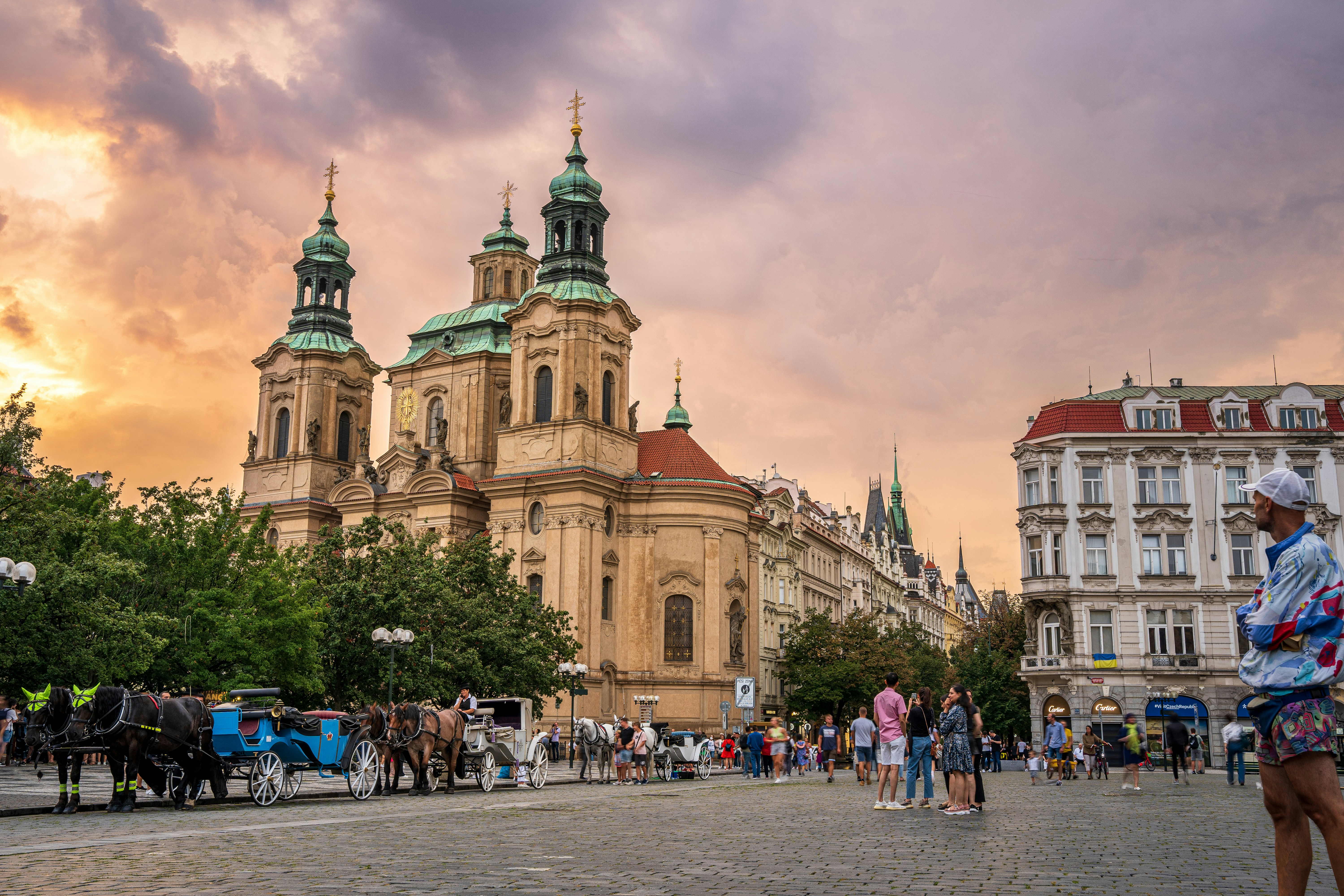 Historic cathedral towers under a vibrant sunset sky with people walking in the plaza.