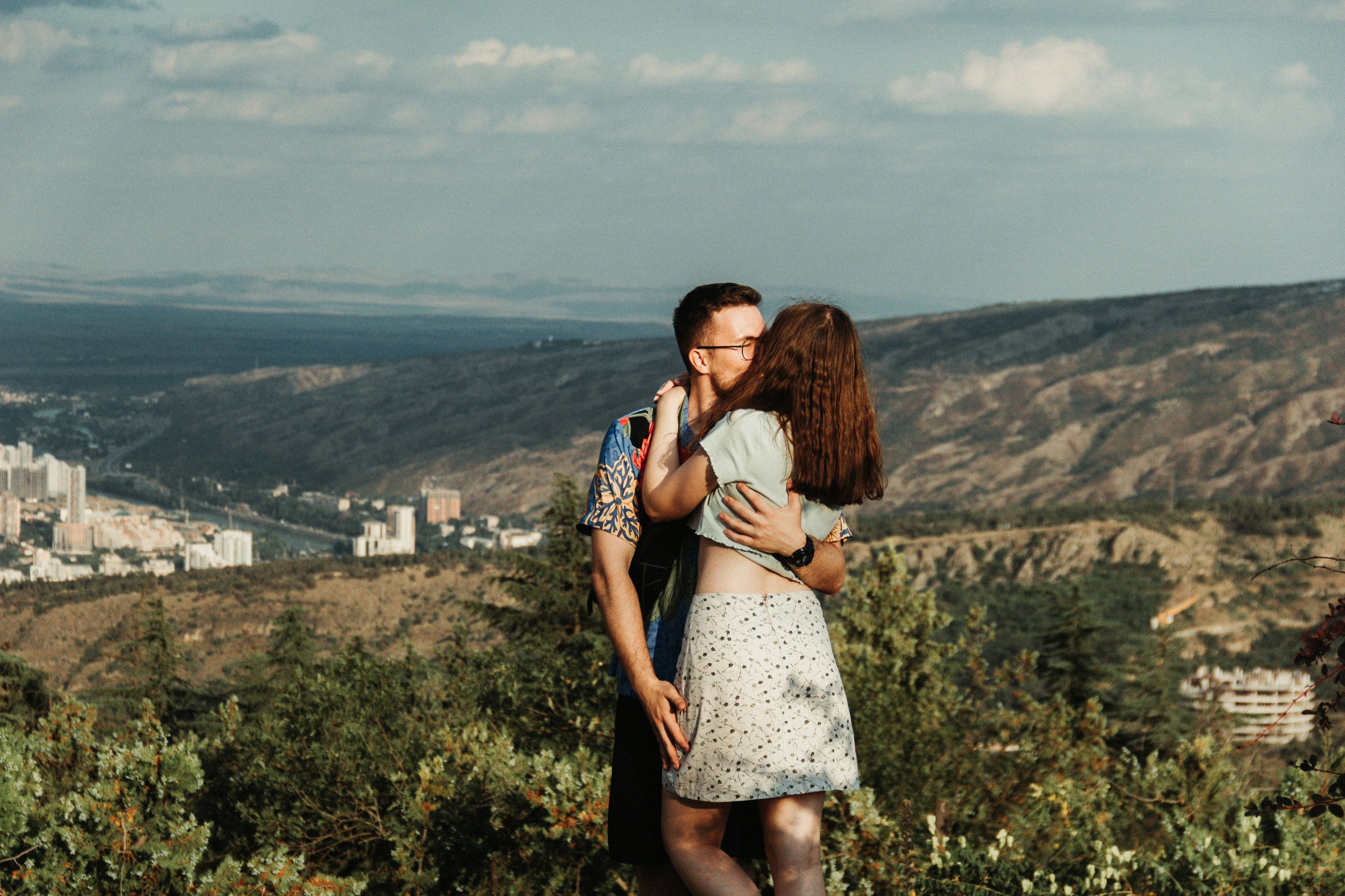Couple embracing on a hillside with a sprawling city and rolling hills in the background.