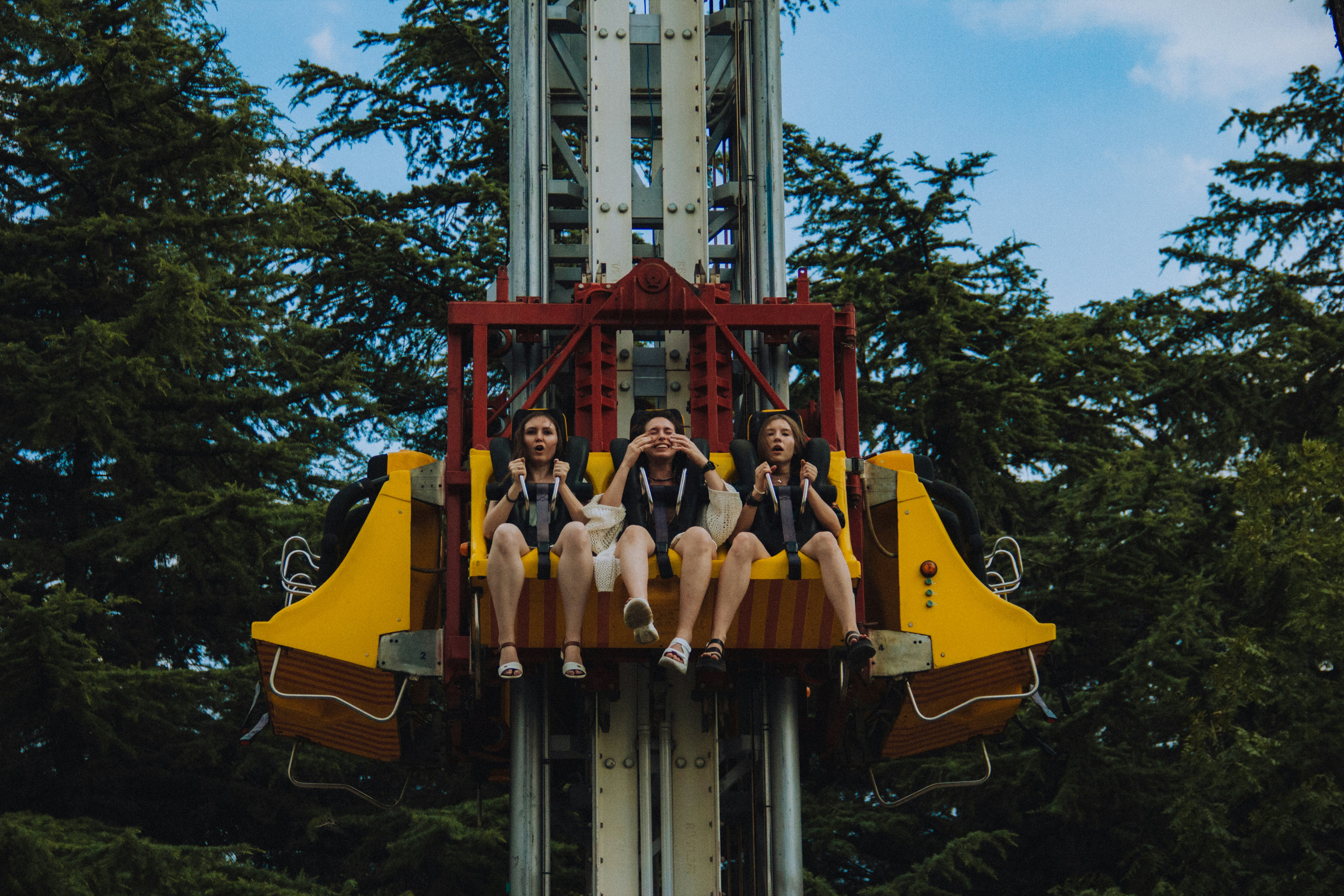 Three riders bracing for a drop on a towering amusement park ride against a backdrop of lush trees.