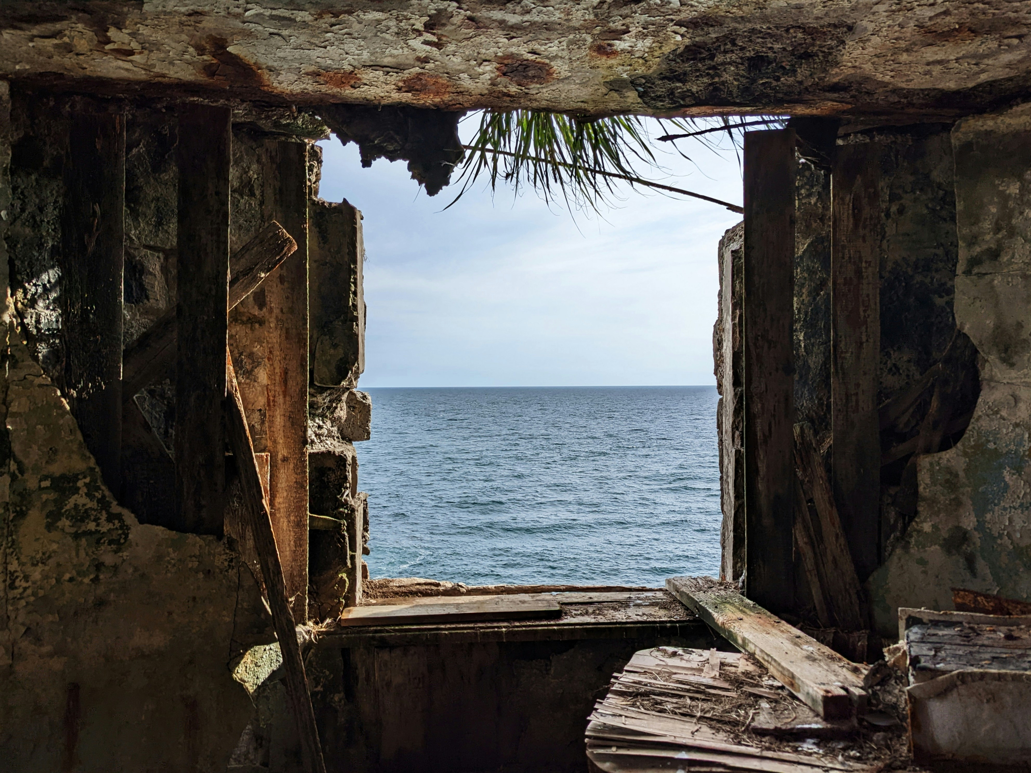 Sea framed by a ruined stone window, with weathered timber and rubble in the foreground. The open frame draws the eye toward the calm blue horizon.