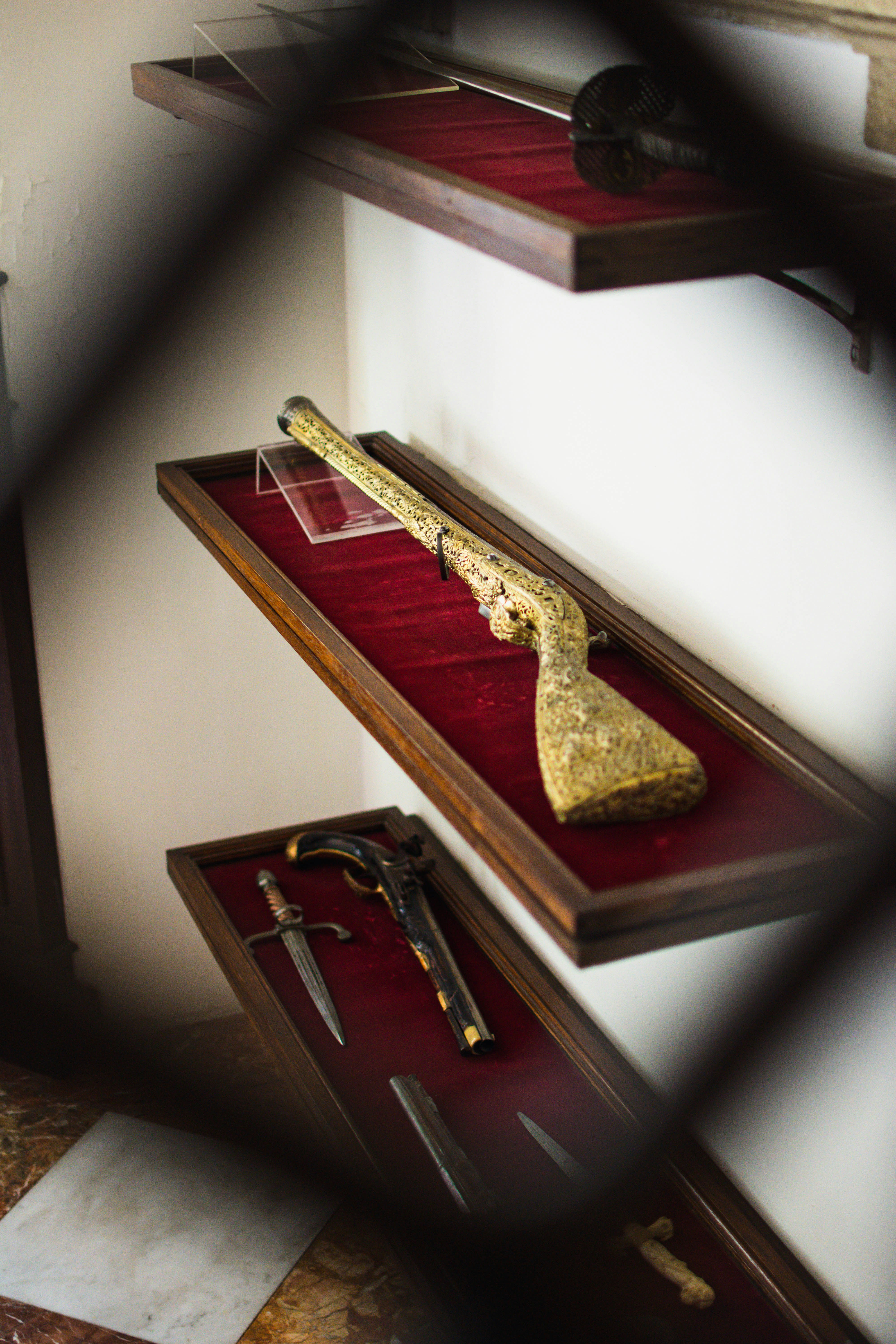 Ornate gold firearm displayed on red velvet inside a glass case, with smaller knives beneath, captured as a museum-style still life.