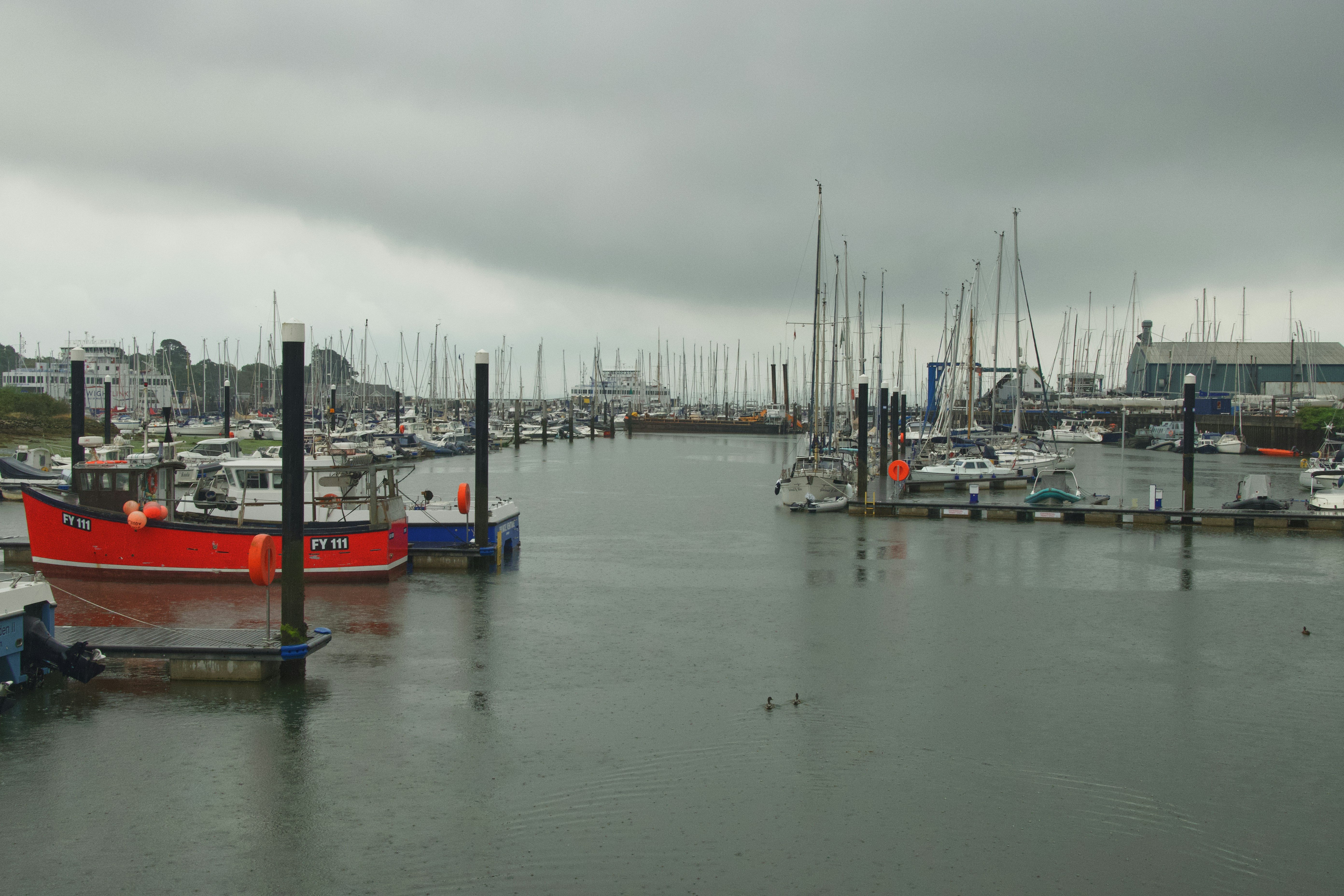Fishing boats and yachts moored in a tranquil harbor under overcast skies, reflecting the calm before the rain. 