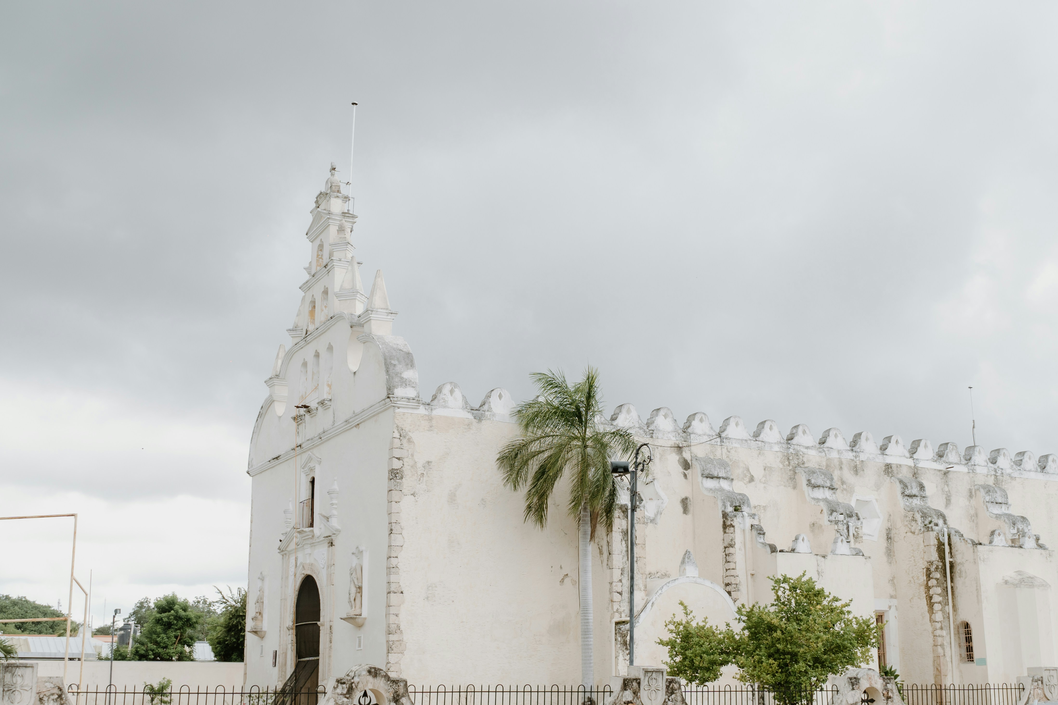 White church with intricate facade stands under a cloudy sky, framed by a palm tree and greenery.