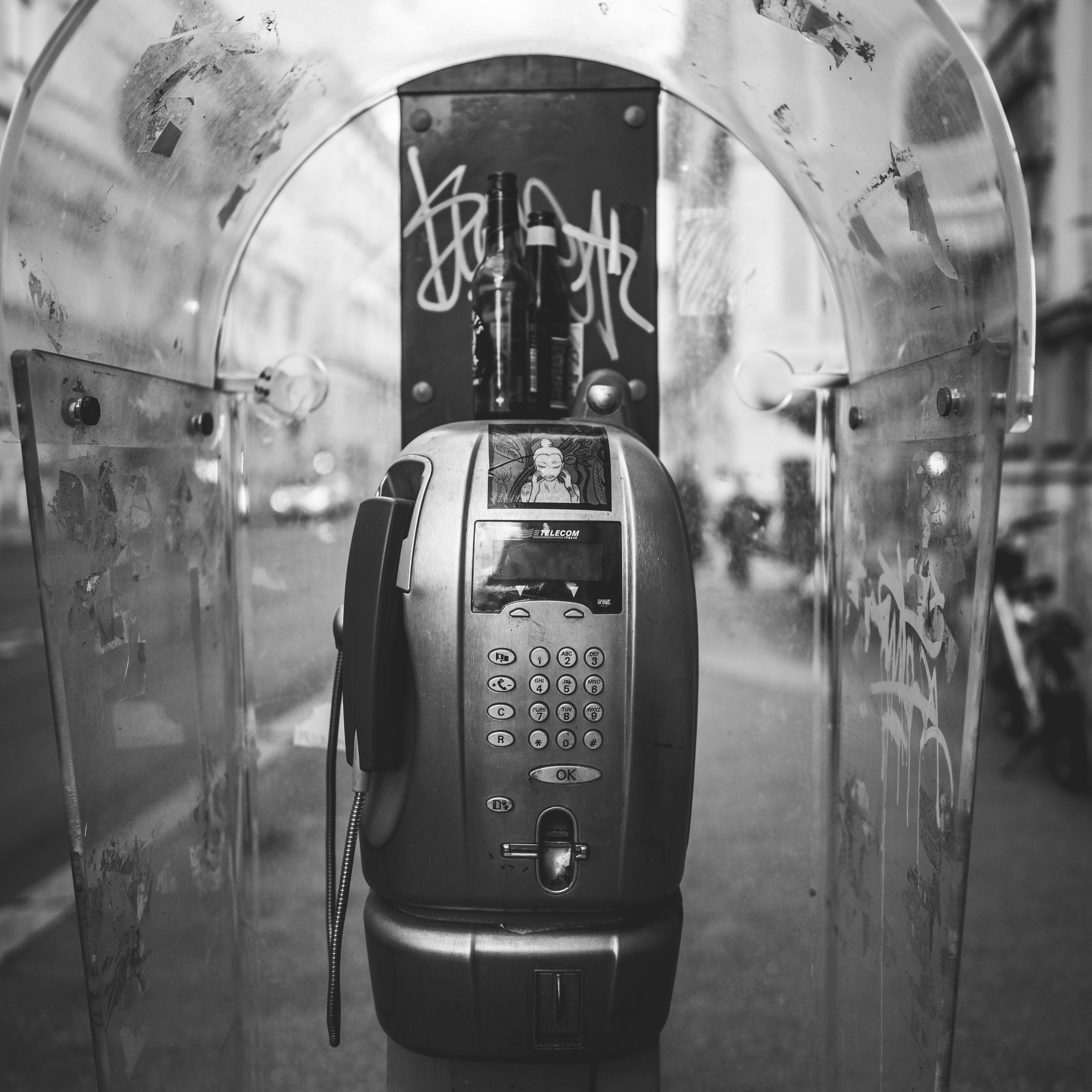 A weathered payphone encased in glass, featuring a reflection of a person, set against a blurred urban backdrop. The scene captures the juxtaposition of past and present.