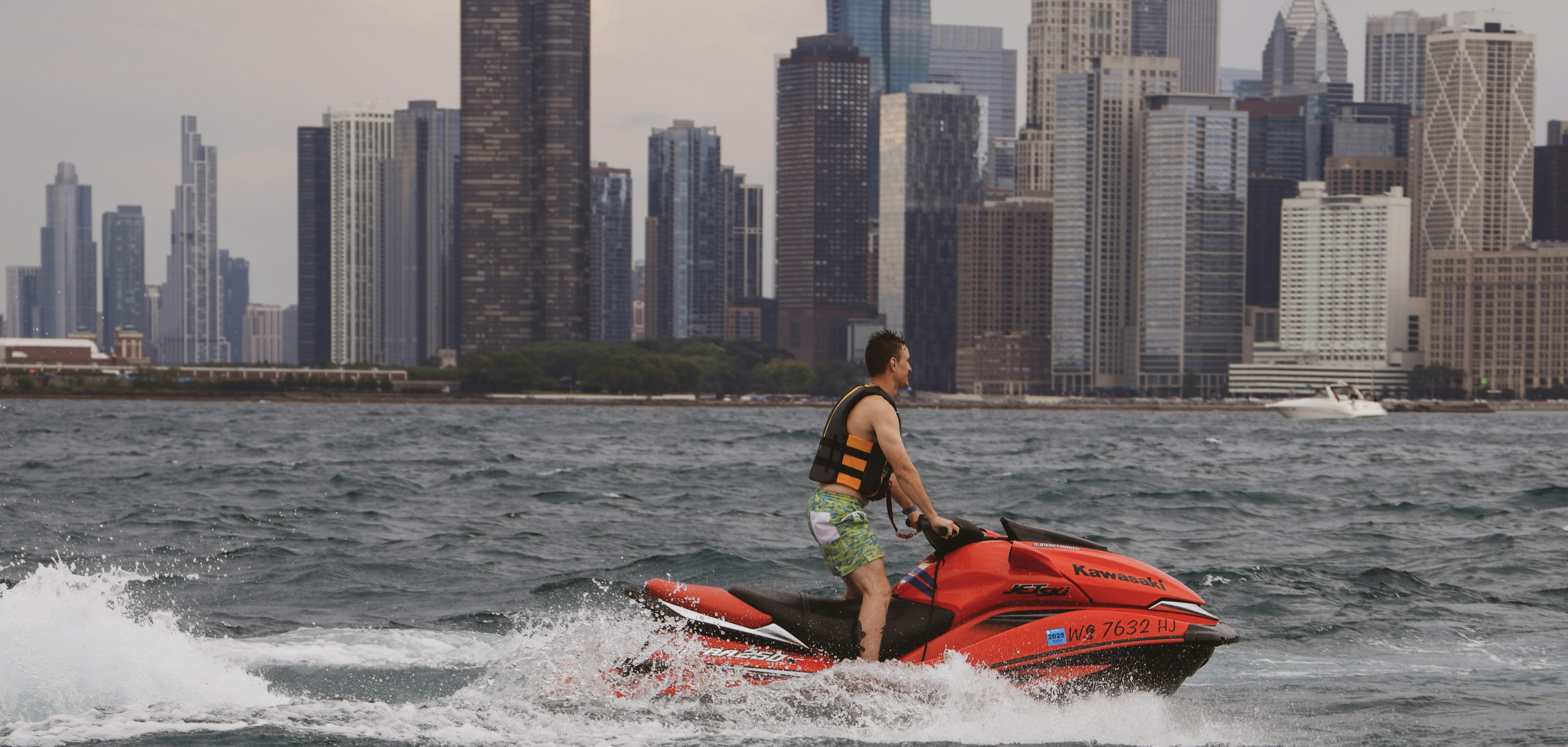 A rider on a bright red jet ski navigates the choppy waters, with a backdrop of towering skyscrapers in a bustling city. The scene captures the thrill of water sports against an urban landscape.