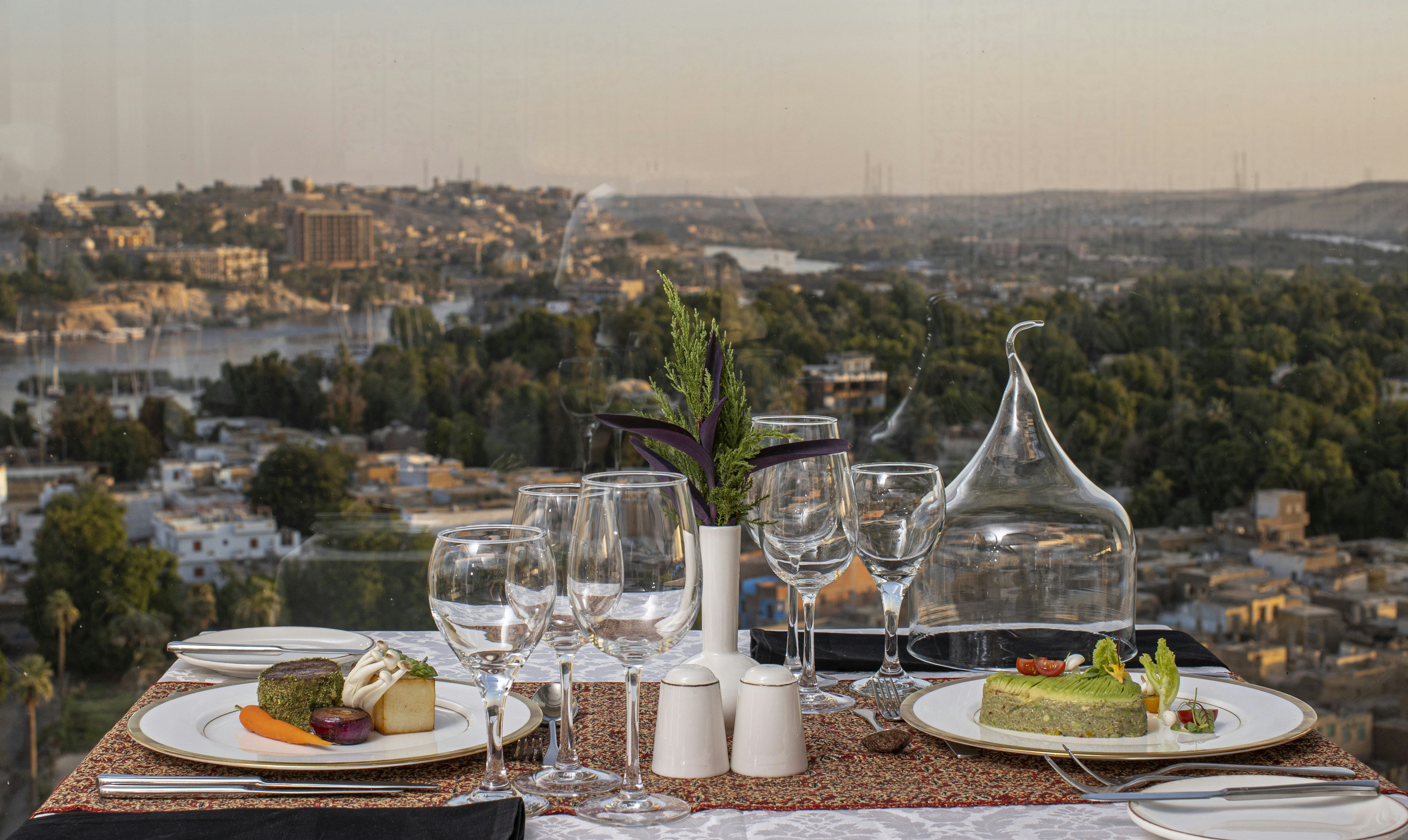 Elegant dining setup featuring gourmet dishes and fine glassware, with a panoramic view of the Nile River and cityscape in the background.