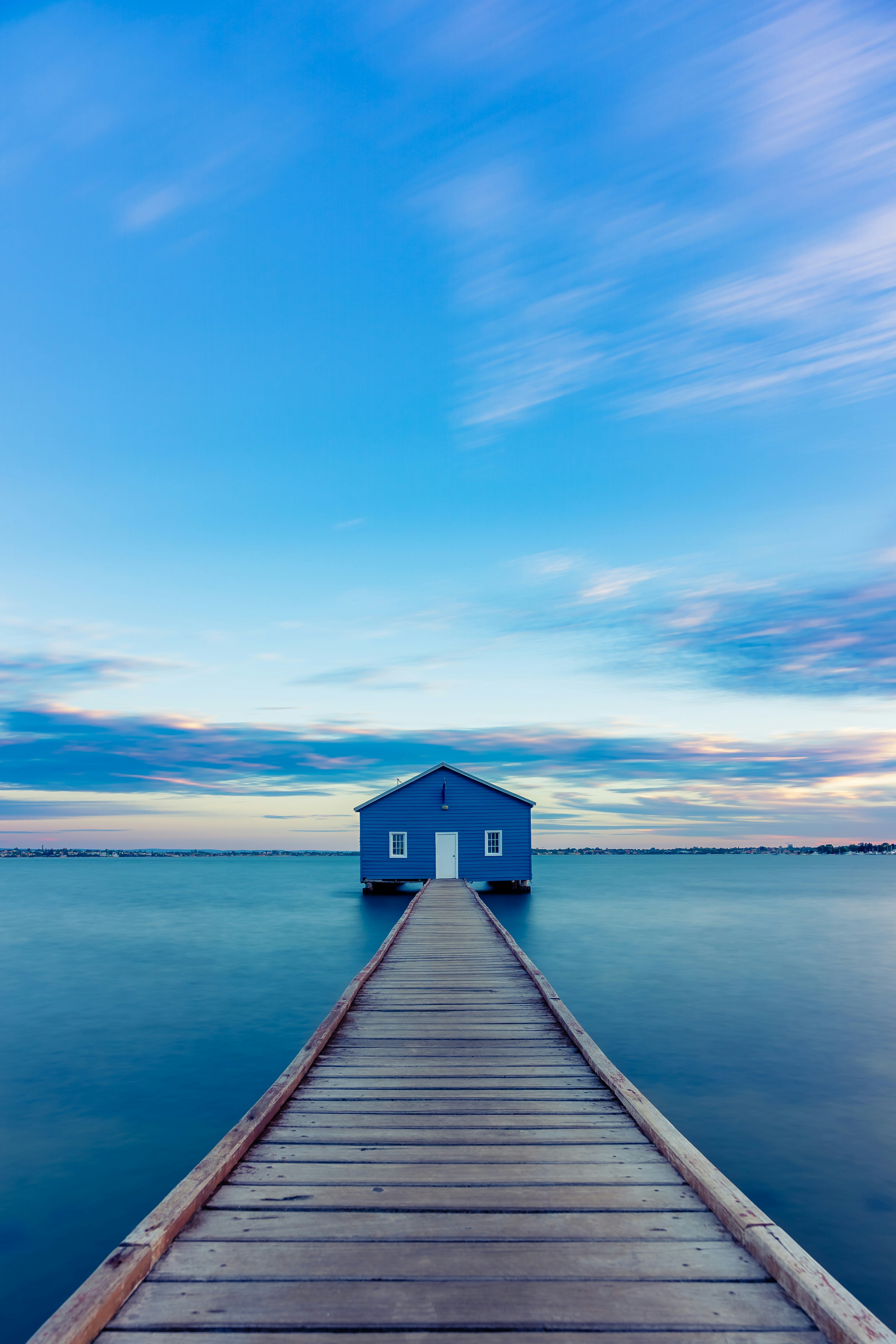 A blue boathouse stands at the end of a wooden pier, reflecting on calm waters under a pastel sky at dusk.