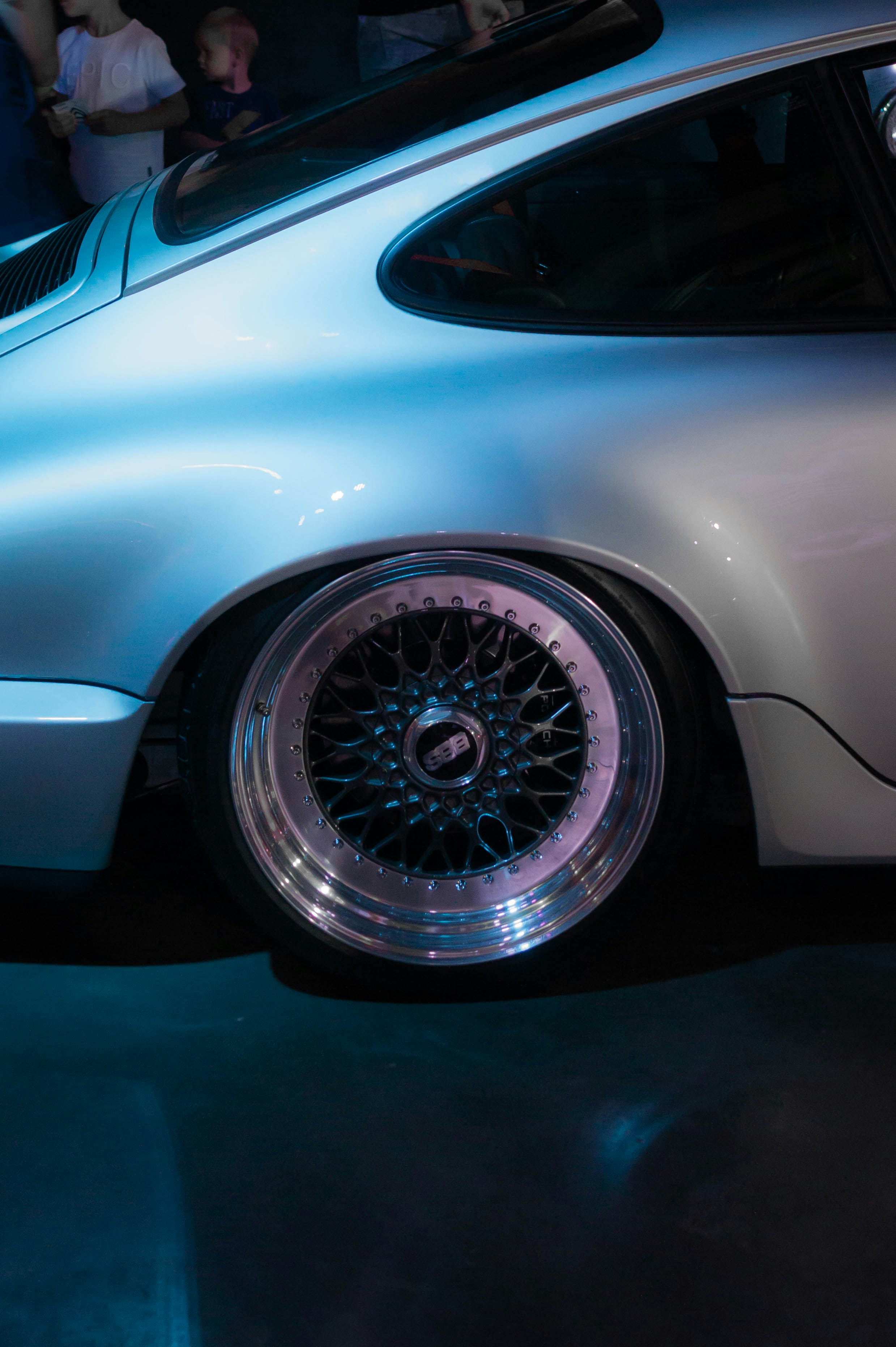 Close-up photograph of a blue-silver sports car wheel with a mesh spoke design, bathed in cool showroom lighting. The image focuses on the wheel hub and polished rims.