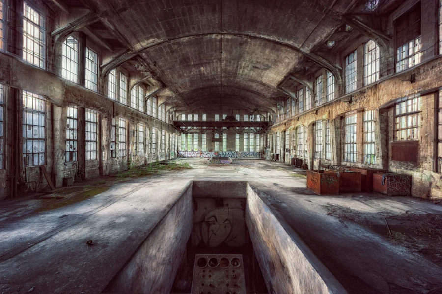 A photograph of an abandoned factory hall featuring a sunken central pit, weathered brick walls, and rows of tall windows streaming diffused light.