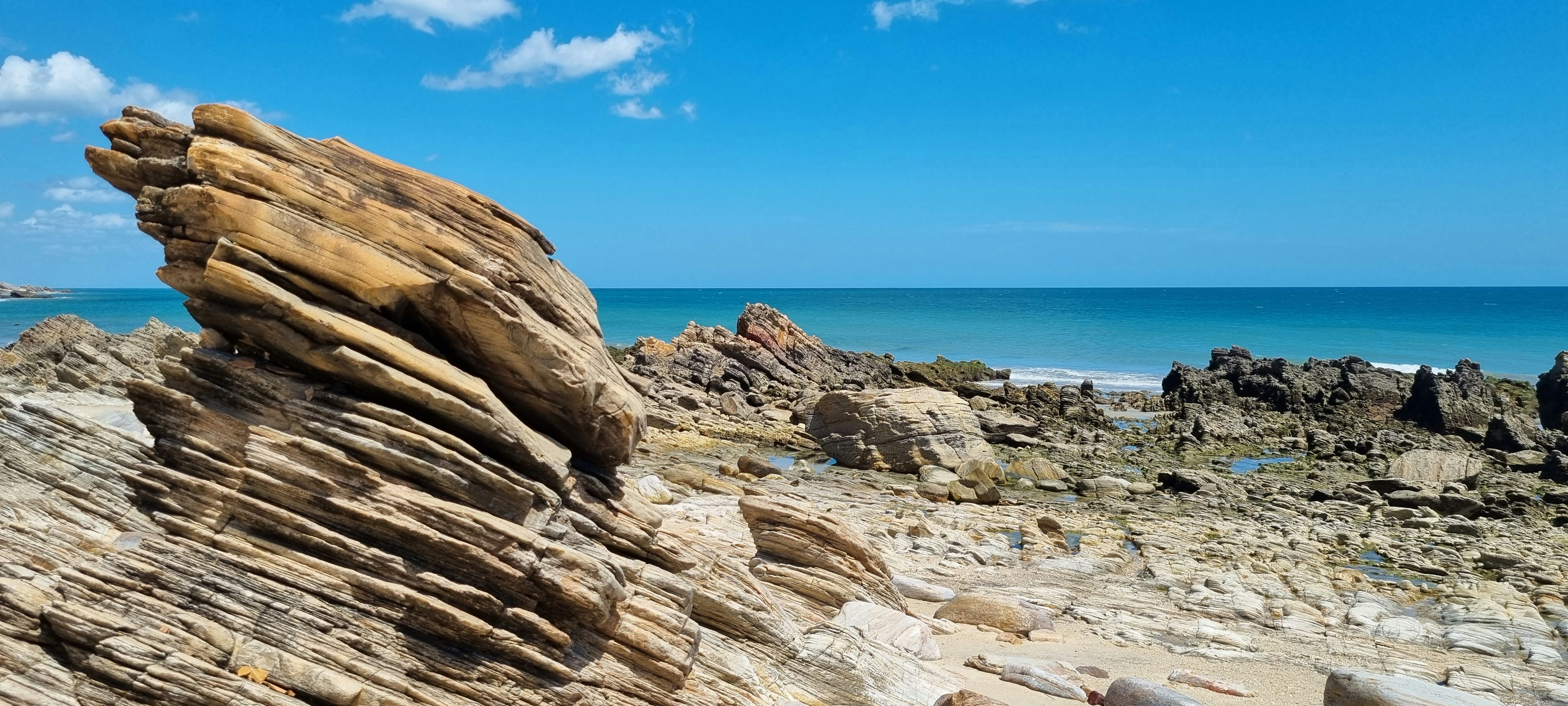 Unique rock formation resembling a sentinel stands prominently against a serene seascape, with gentle waves lapping at the shore. The clear blue sky enhances the coastal scenery.