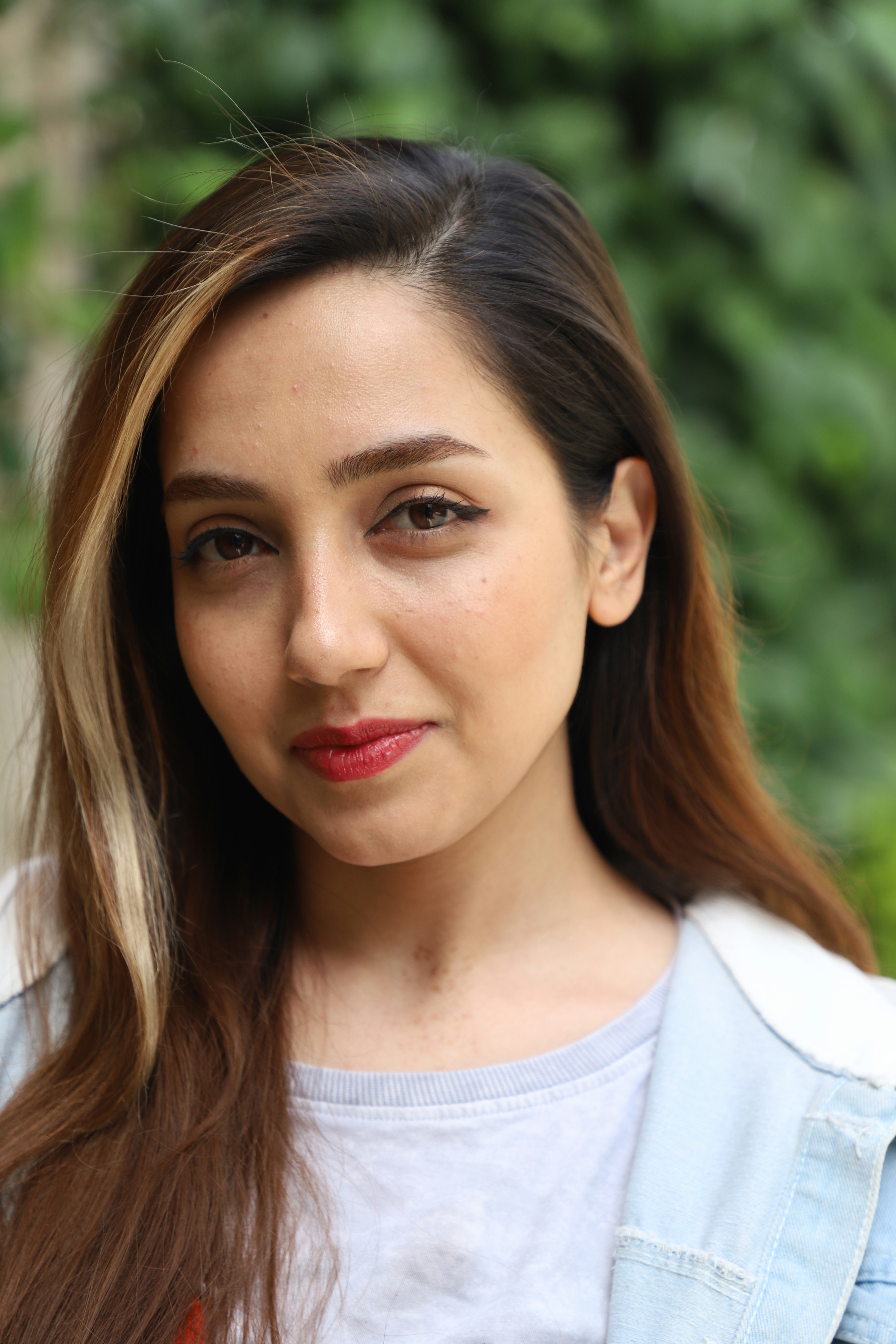 Close-up portrait of a woman with long hair and subtle makeup, set against a lush green background.