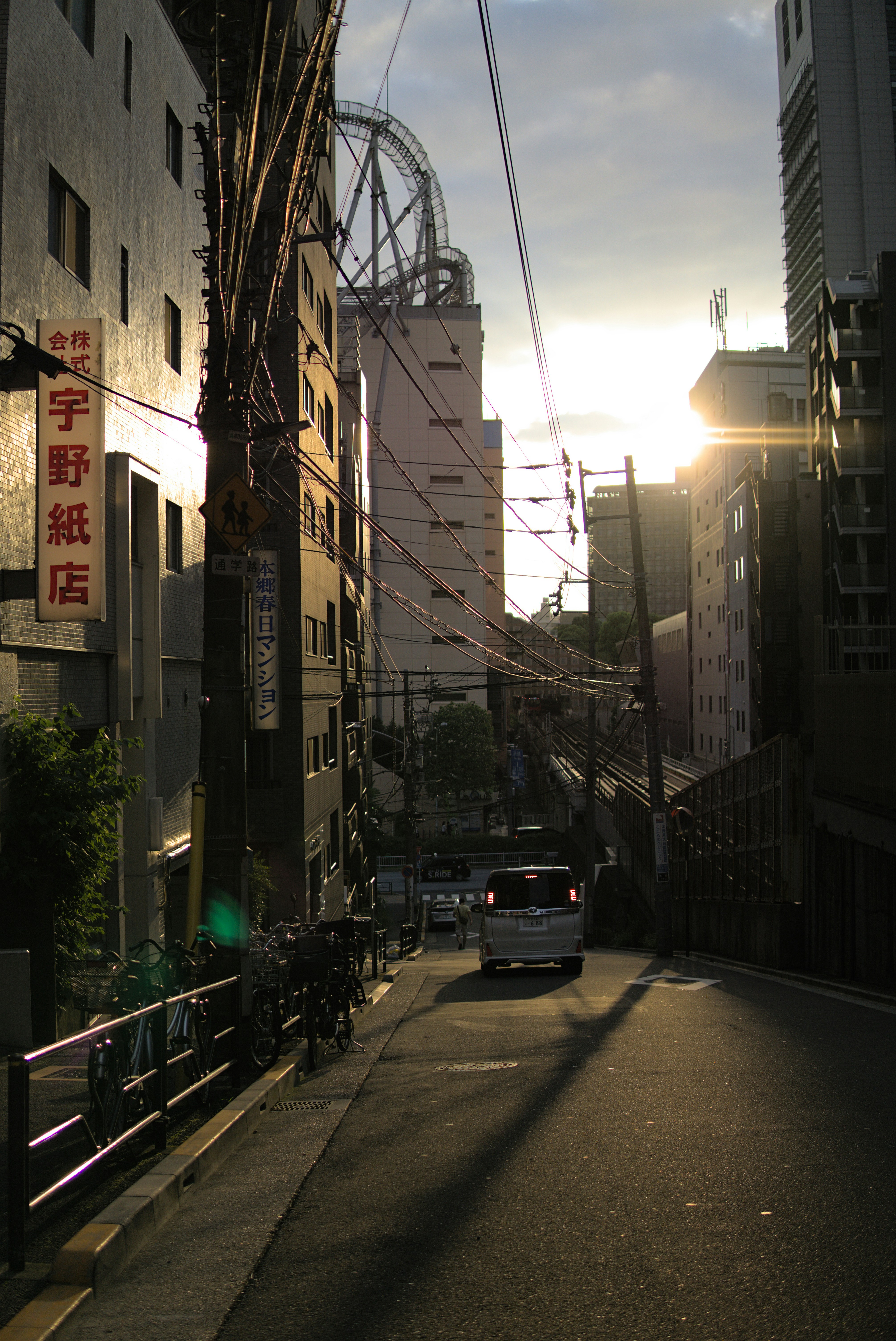 Sunset illuminating a narrow urban street, with buildings and power lines creating a dynamic composition. A vehicle navigates the incline as shadows stretch across the pavement.