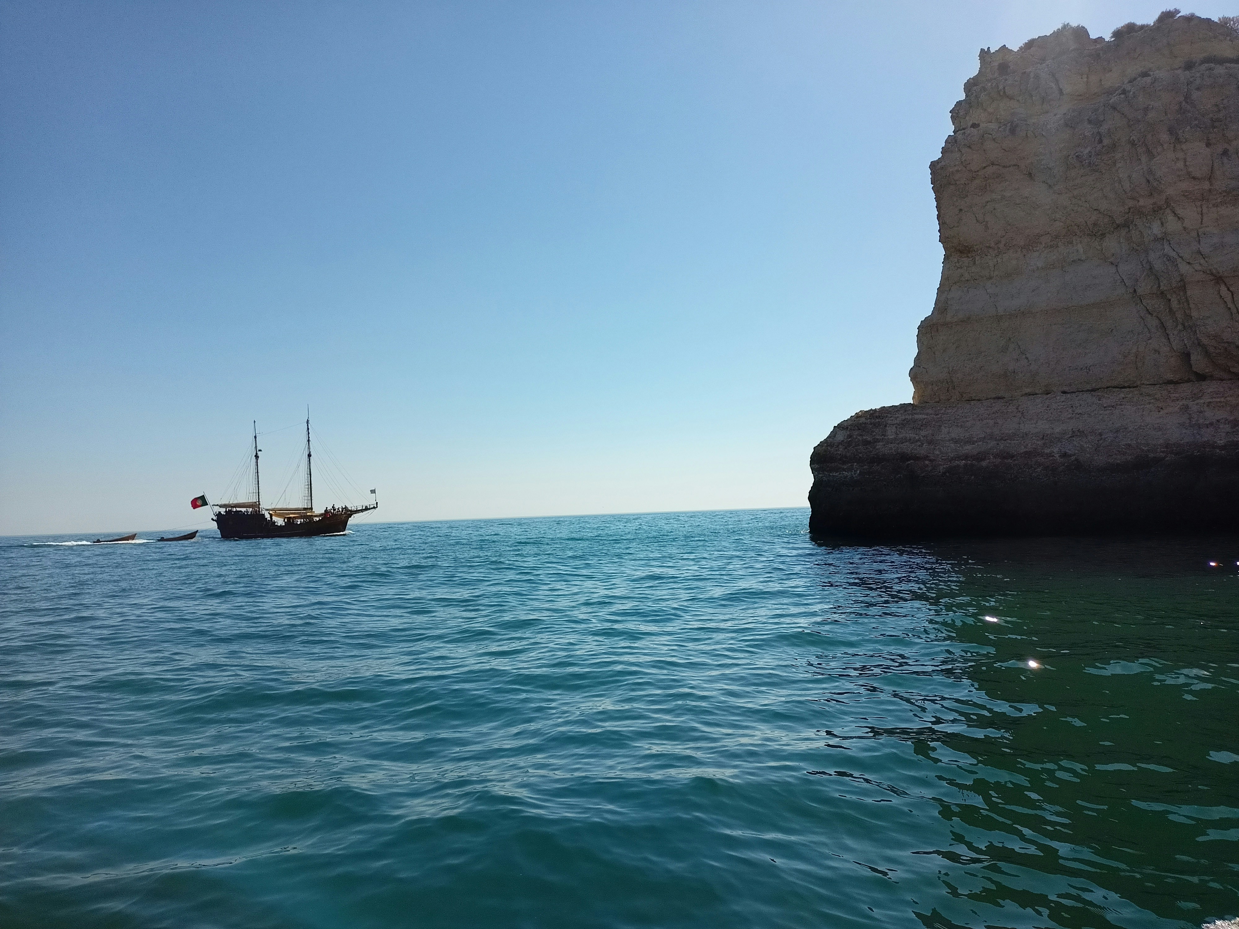 Sailing ship on calm blue sea near a towering cliffside, bathed in bright daylight.